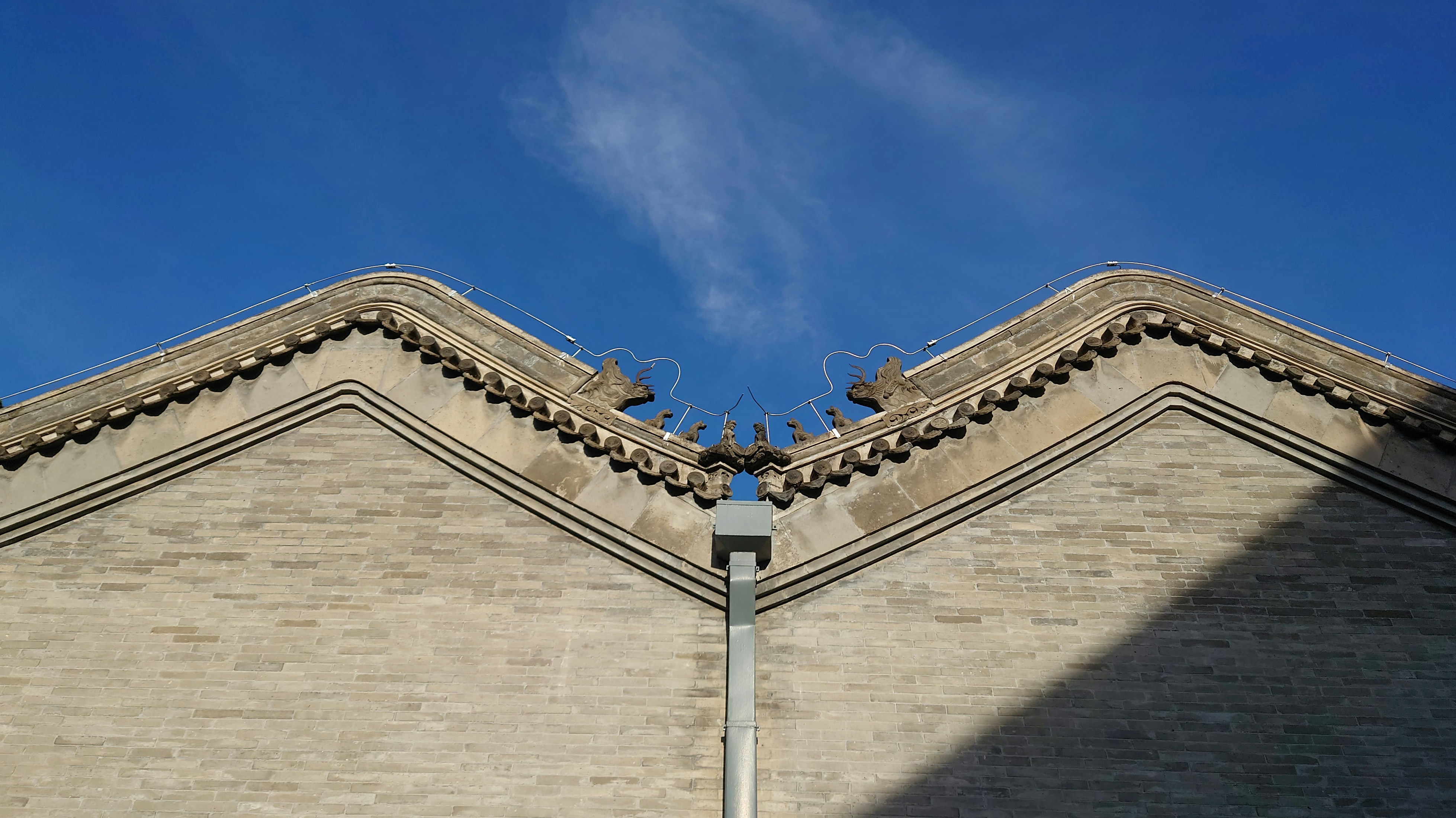 Symmetrical brick roof arches converge at the center against a clear blue sky, with a shadow sweeping across one side and a small metal fixture at the ridge.