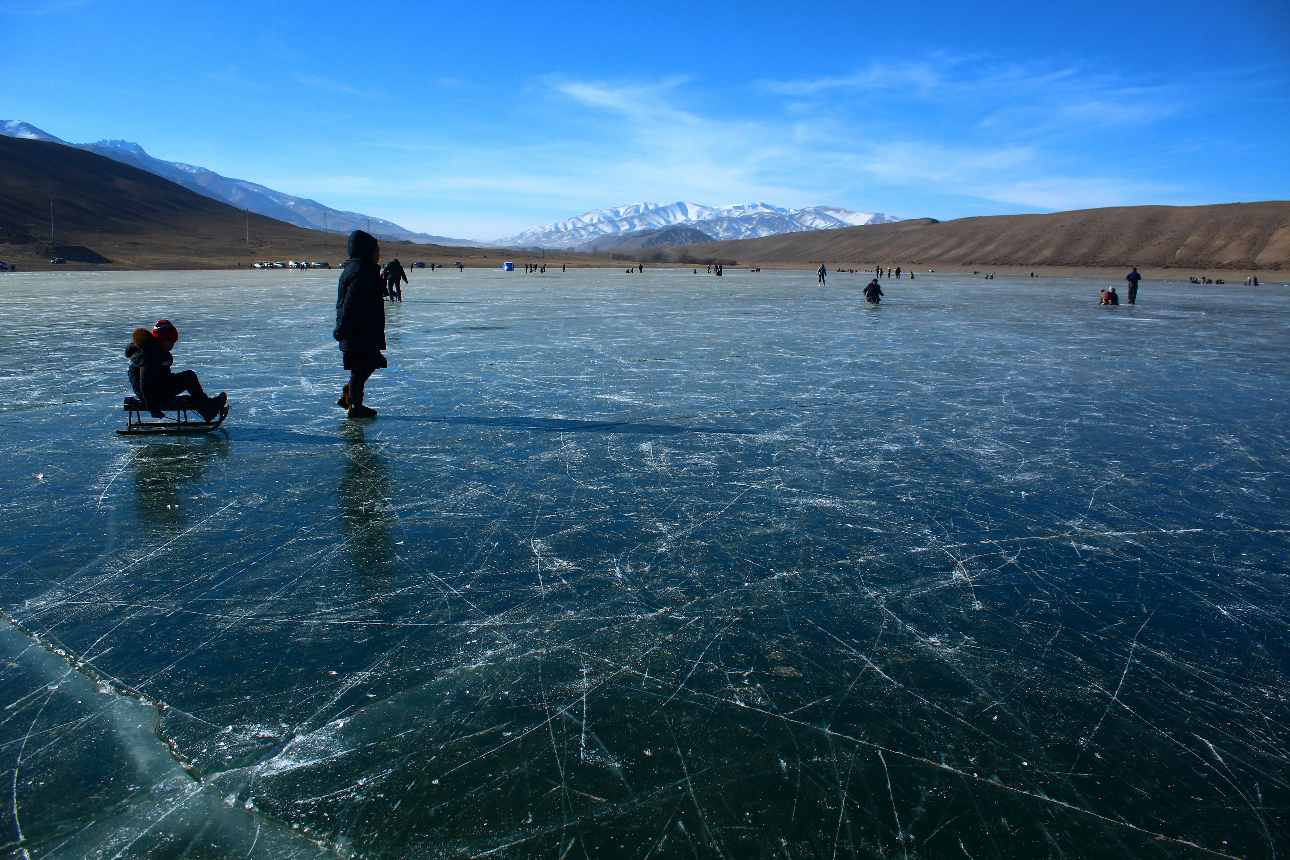 People walking and skating on a vast frozen lake under a clear blue sky, surrounded by distant mountains.