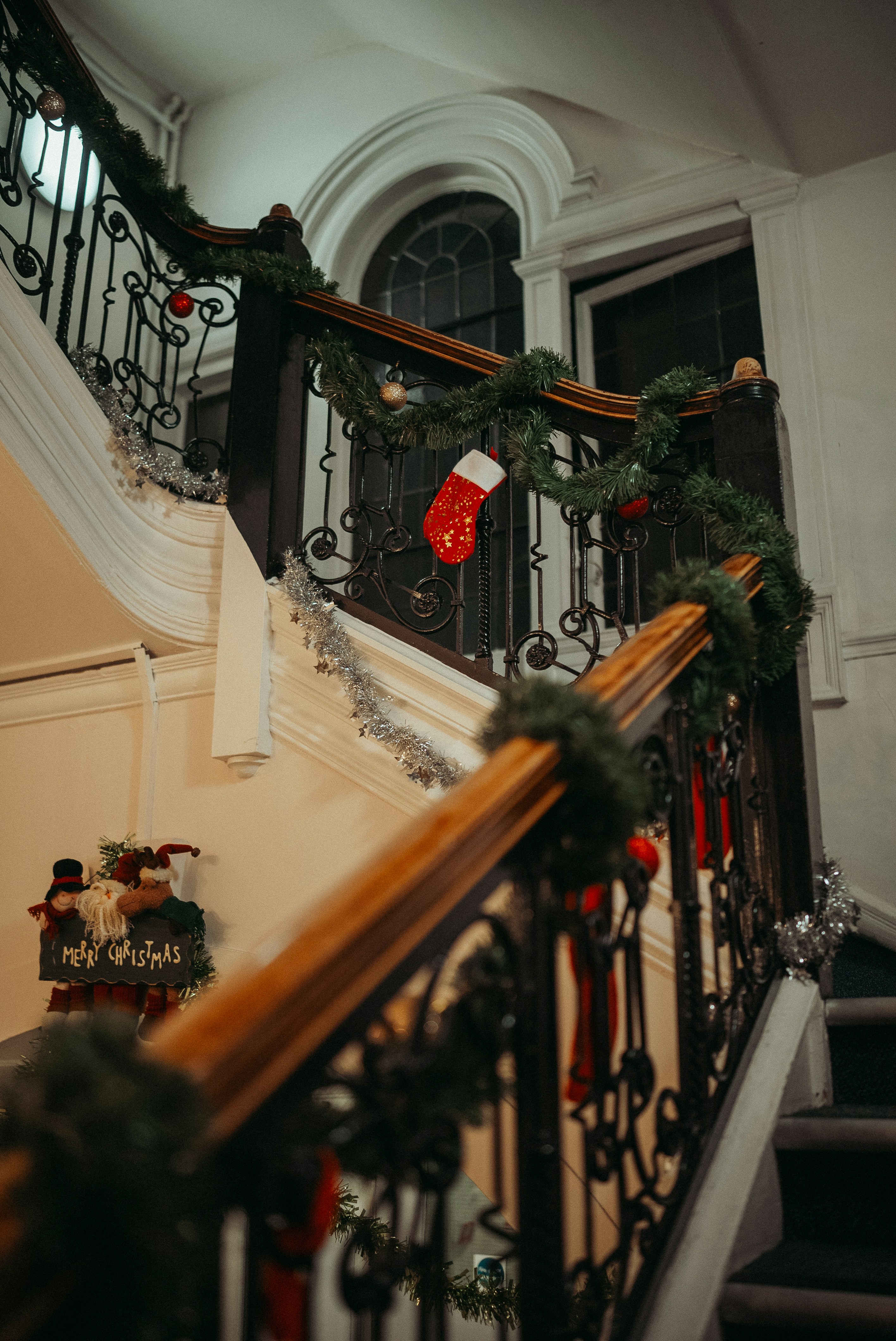Festively decorated staircase with garlands, lights, and a red stocking hung from an ornate railing, with a small 'Merry Christmas' sign at the lower landing.
