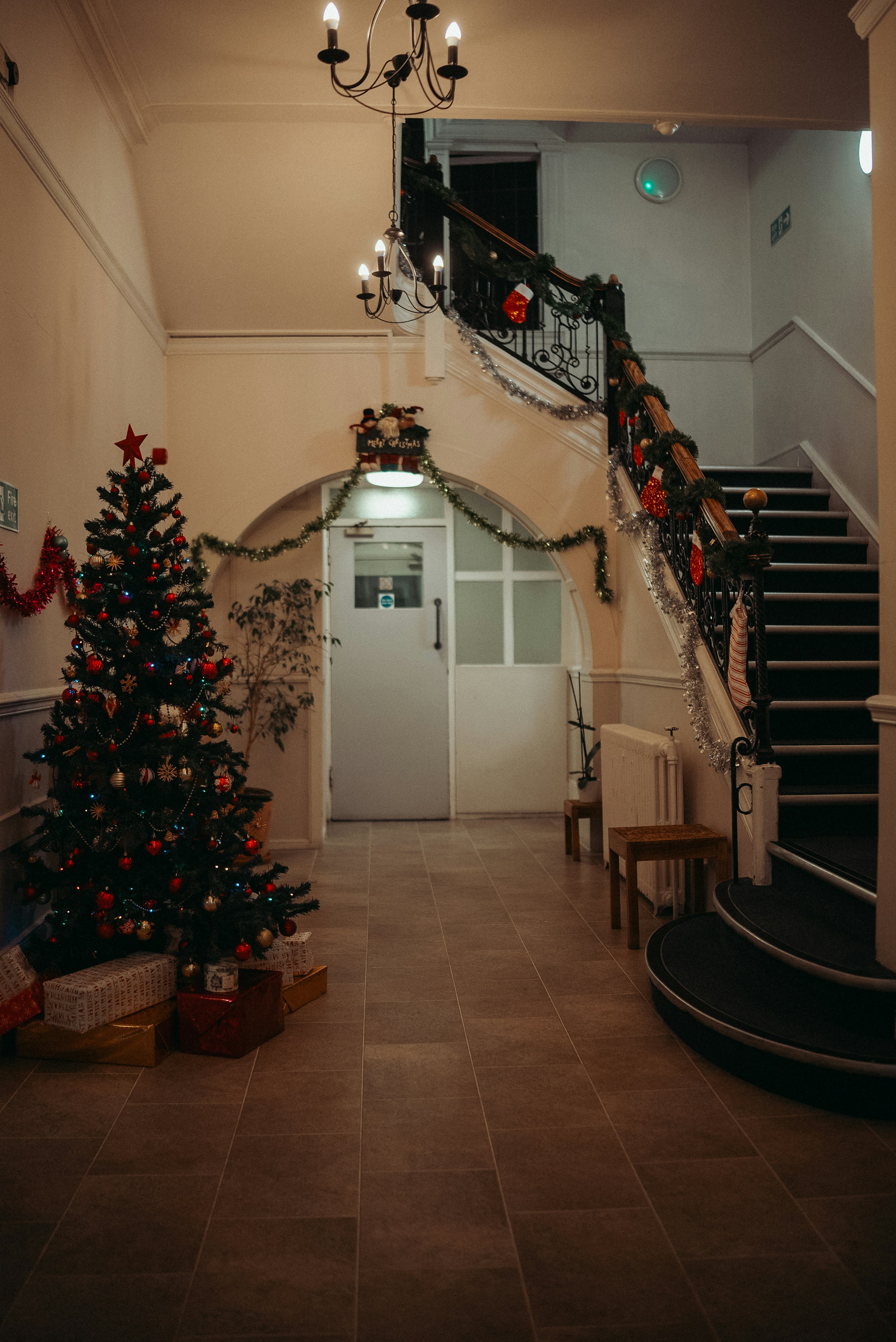 Warmly lit lobby scene with a decorated Christmas tree on the left and a garland-draped staircase on the right, set against a tiled floor.