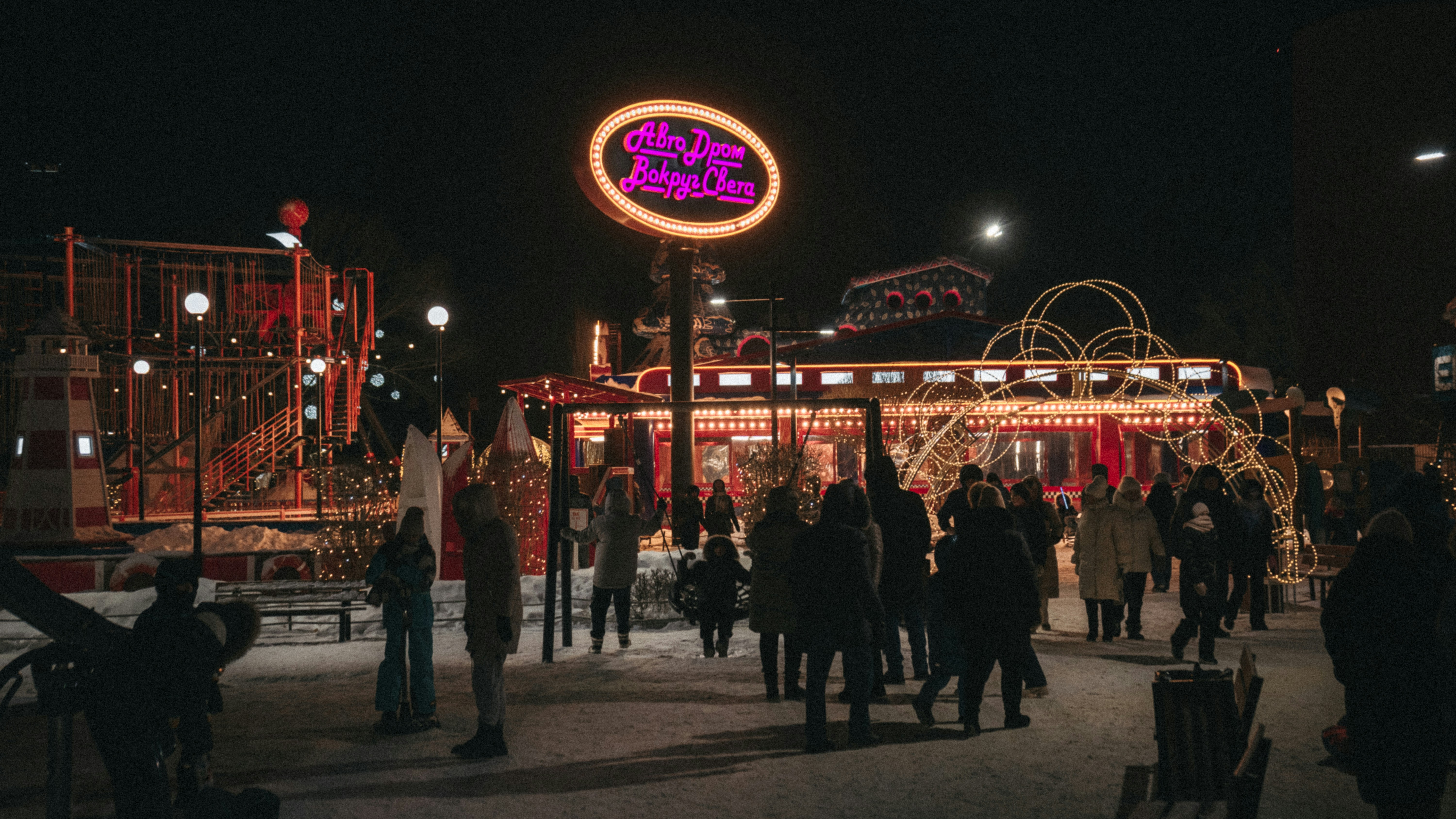 Crowd gathers at a brightly lit amusement park at night, with neon signs and rides in the background.