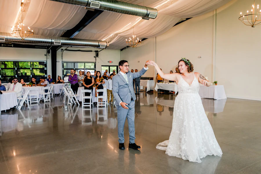Bride and groom dance in the center of a spacious wedding reception hall while guests watch from the tables in the background.