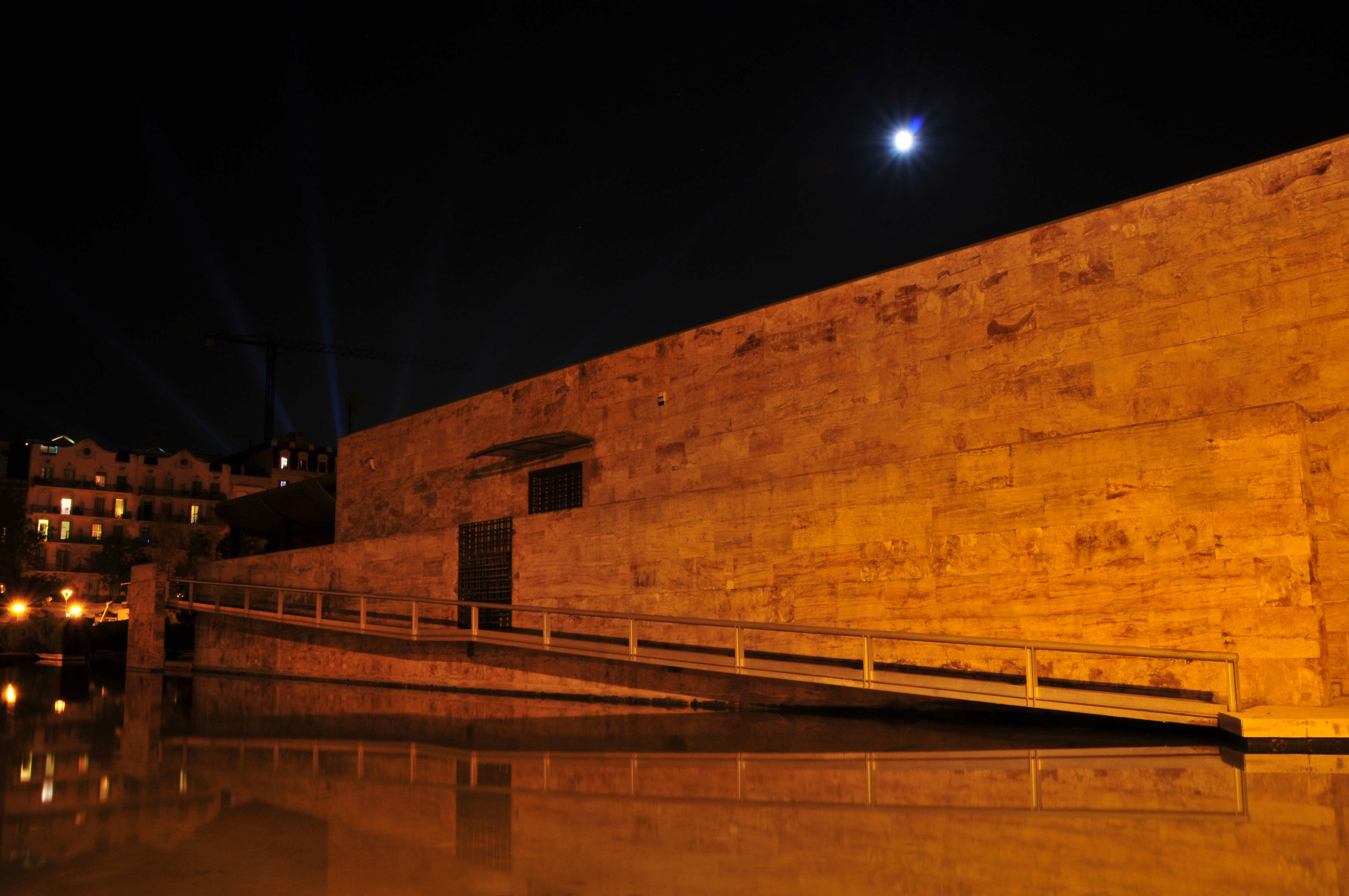 A modern architectural structure illuminated by warm lights, with a serene reflection in the water under a bright moon.