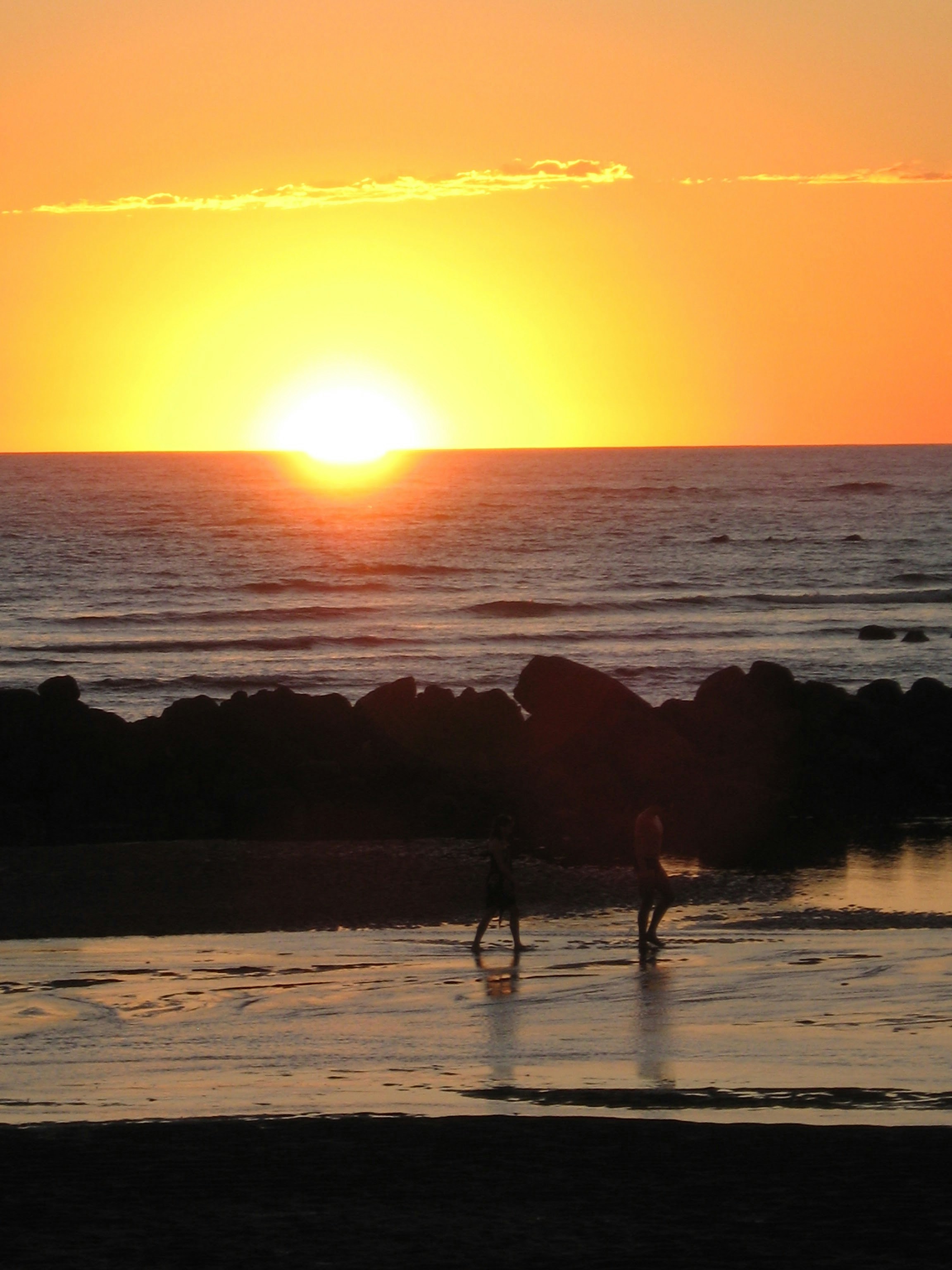 Silhouetted figures walk along the shoreline as the sun sets over the ocean, casting a warm glow on the water.