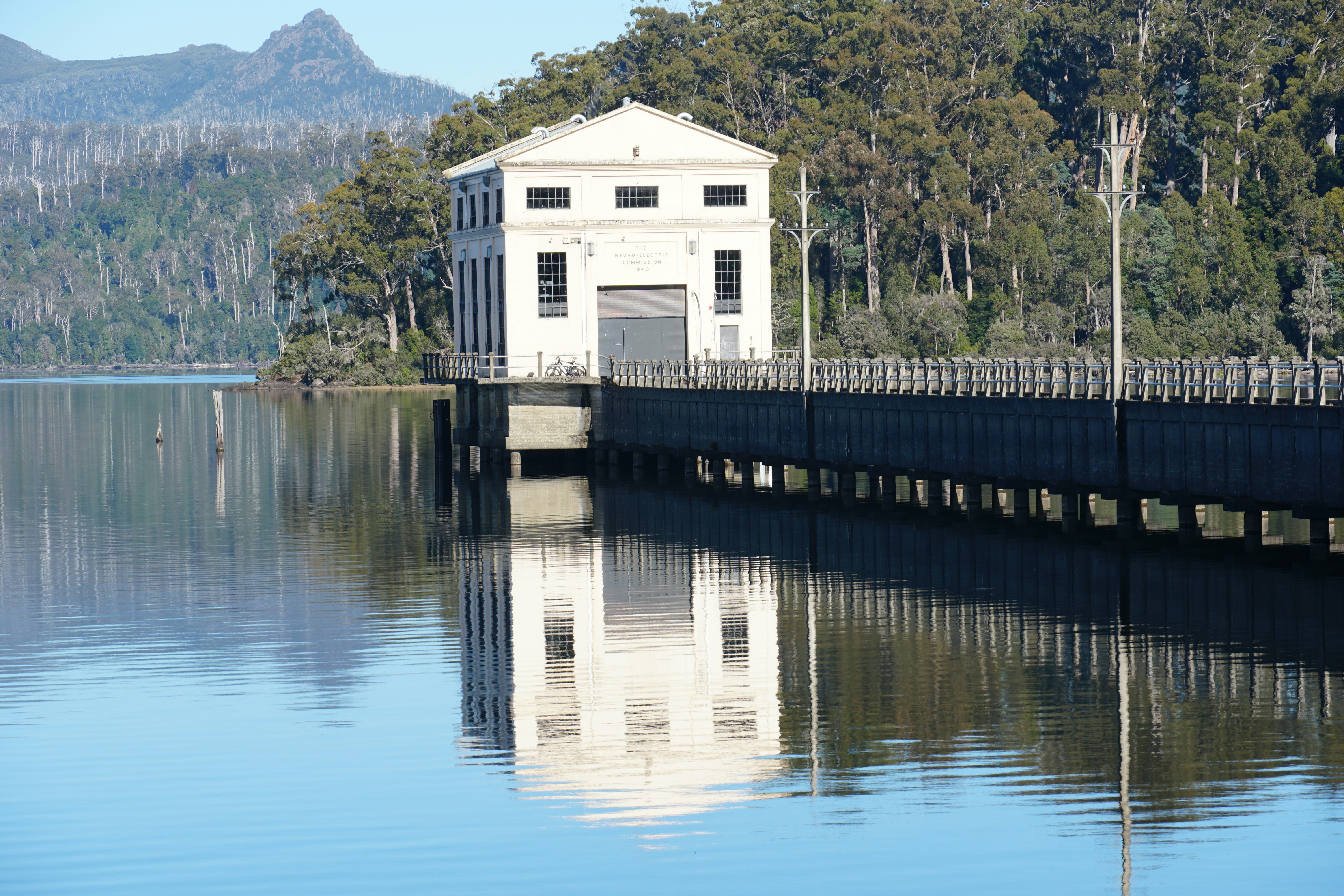 Historic building on a pier reflects in the calm waters of Lake St Clair, surrounded by dense forest and distant mountains.