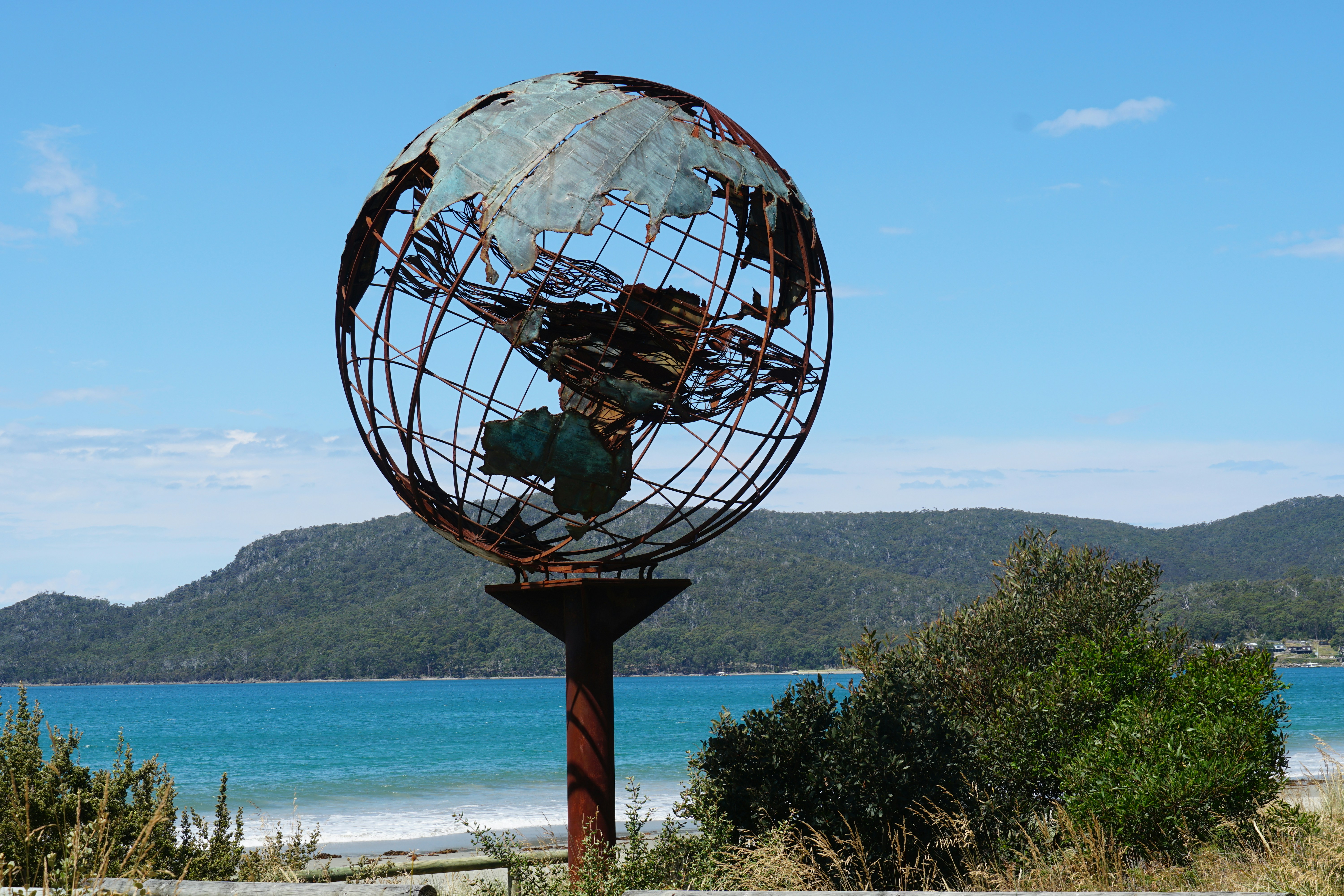 This captivating image features a weathered metal globe sculpture set against a stunning coastal backdrop. The globe's rustic textures contrast beautifully with the vivid blues of the ocean and sky, while lush greenery frames the scene. The interplay of natural and man-made elements, along with the bright, natural lighting, creates a visually striking composition that evokes a sense of global wanderlust.