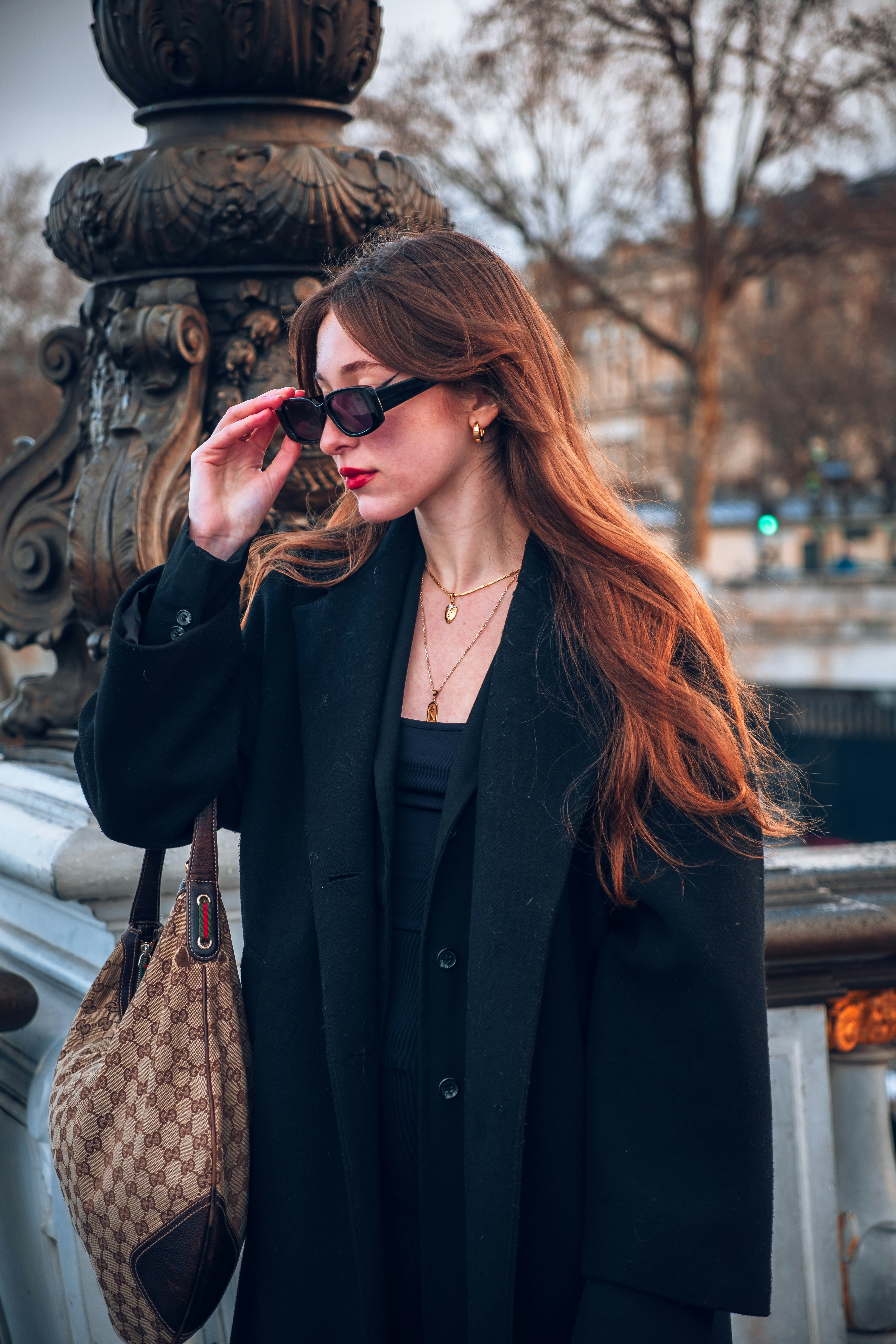 A stylish woman in a black coat adjusts sunglasses beside an ornate lamppost along a riverfront, carrying a patterned handbag. This photograph captures a moody urban moment.