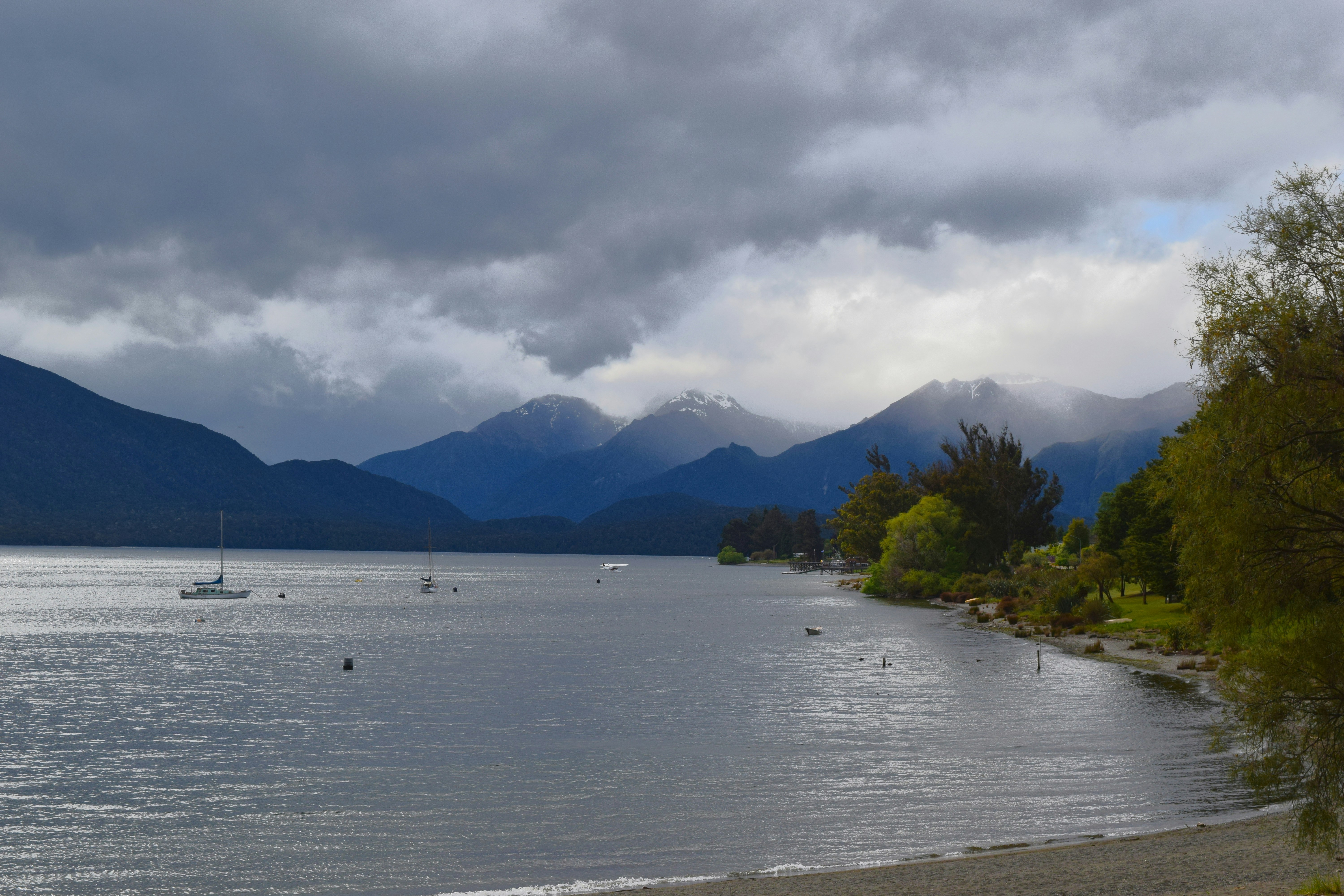 Cloud-covered mountains rise above a tranquil lake shoreline with boats and lush greenery.