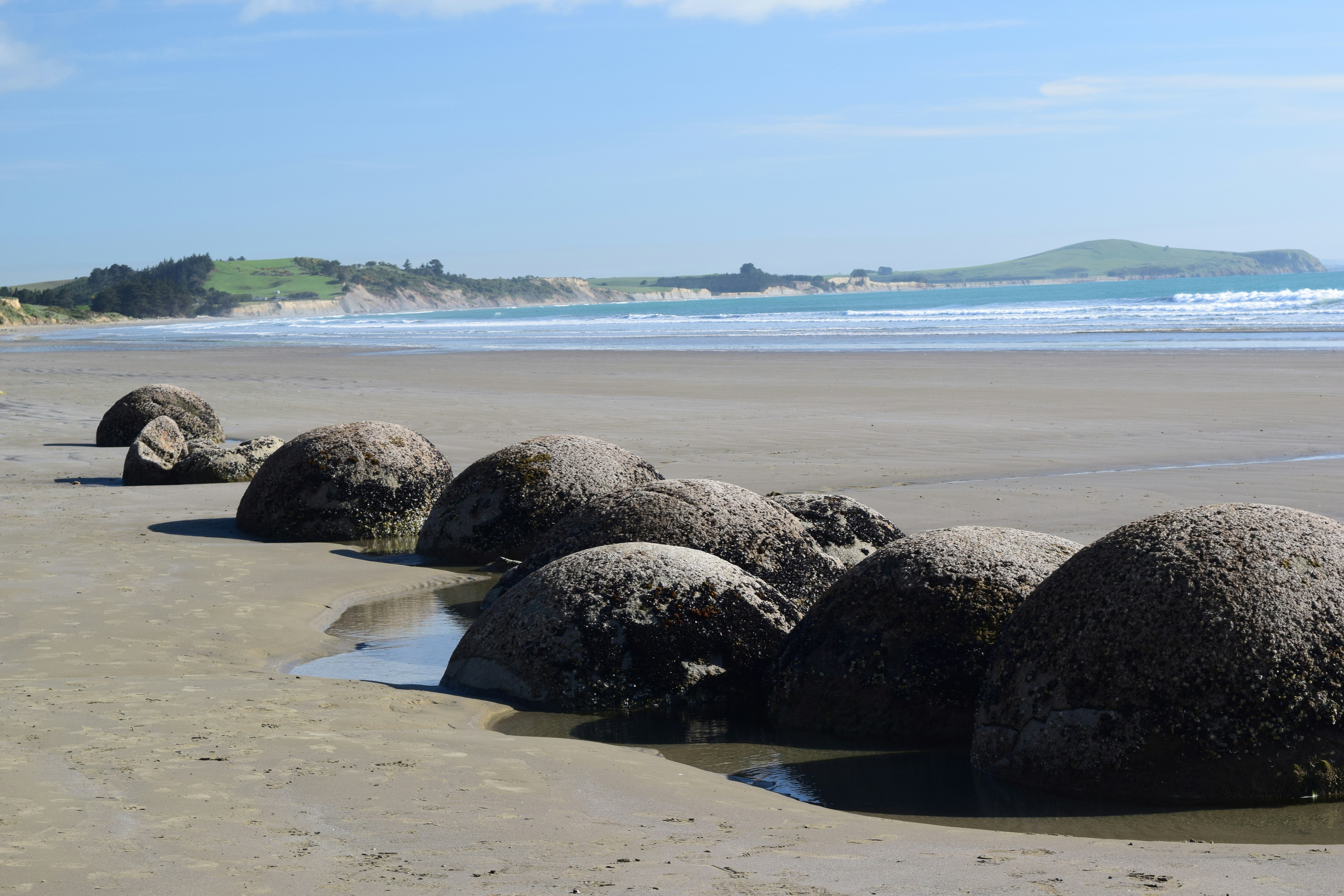 Cluster of large, rounded rocks along a sandy beach with gentle waves and distant headlands under a clear sky.