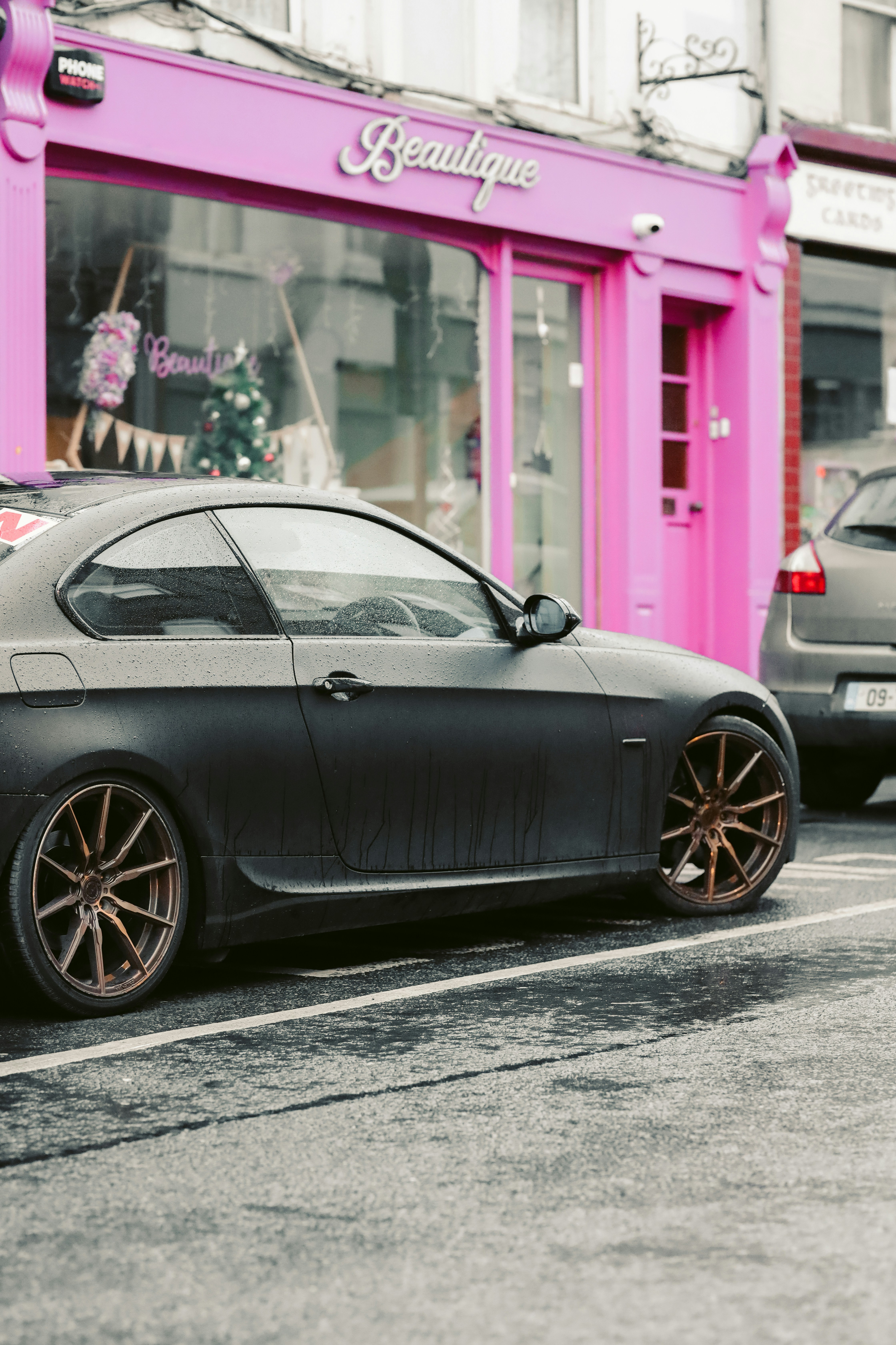 Matte Black Coupe Beside a Neon Pink Boutique on a Rainy City Street