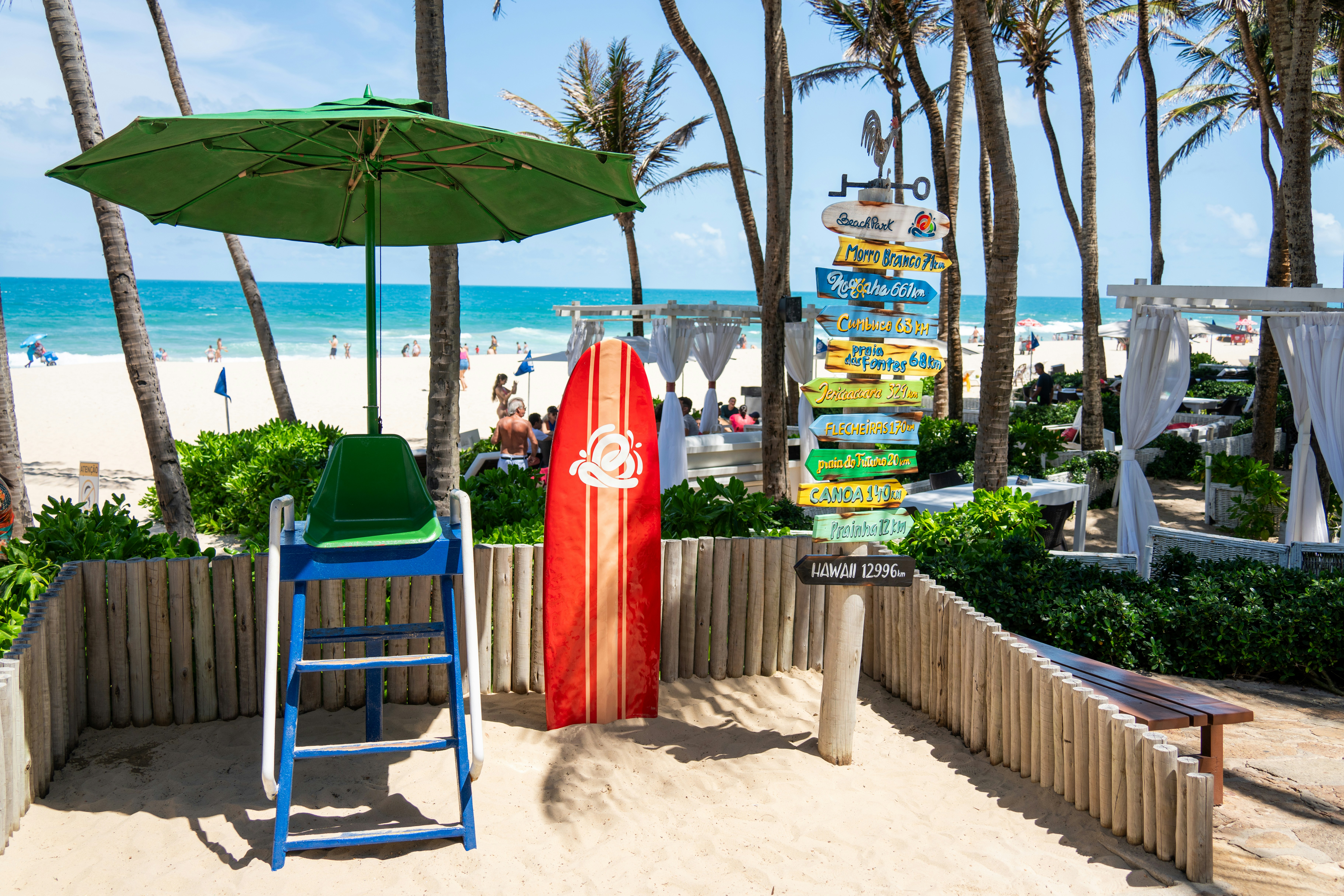 Red surfboard and lifeguard stand under an umbrella on a sandy beach with palm trees and ocean in the background.