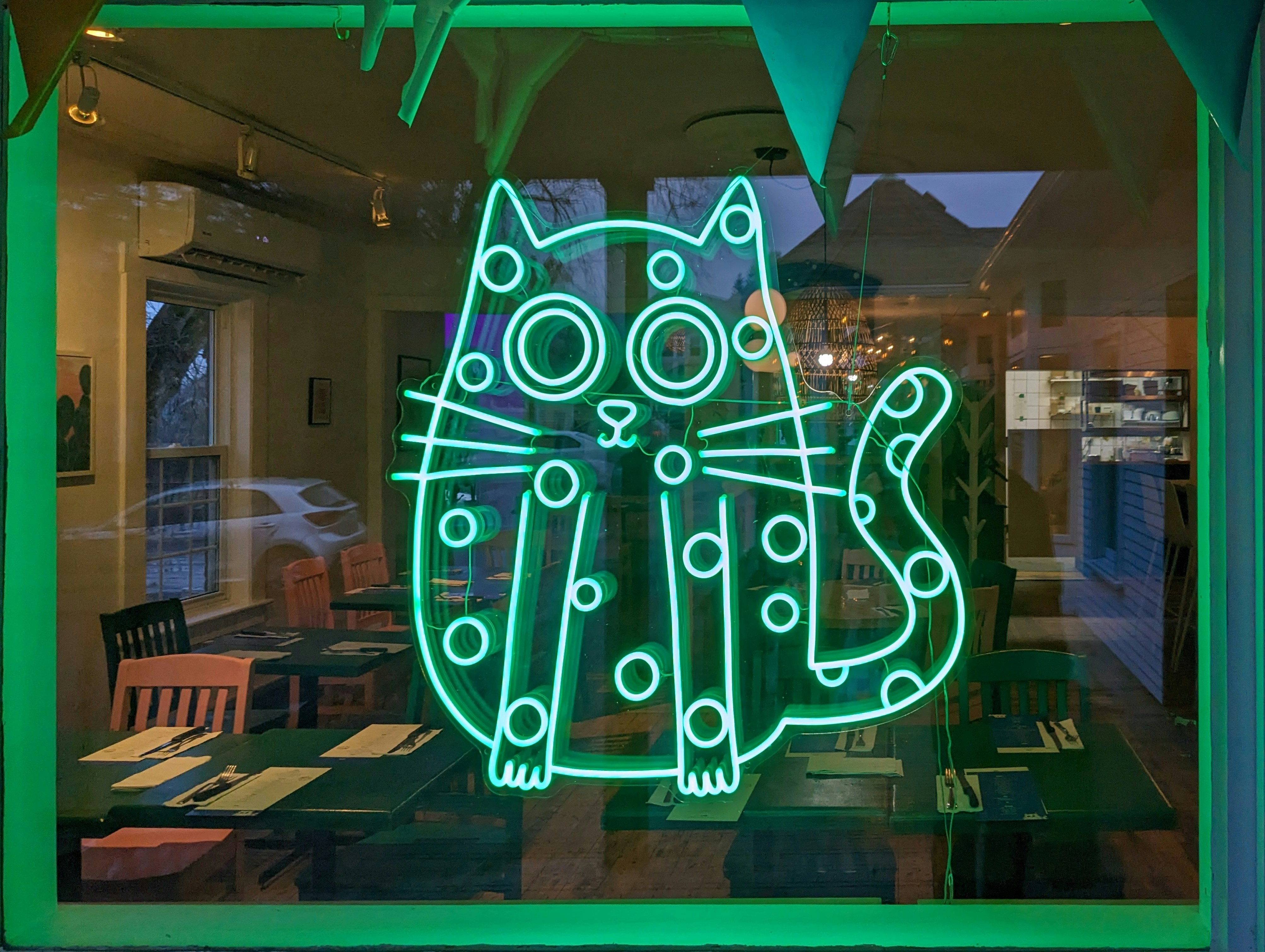 Green neon cat sign glowing in a restaurant window with tables and chairs visible inside.