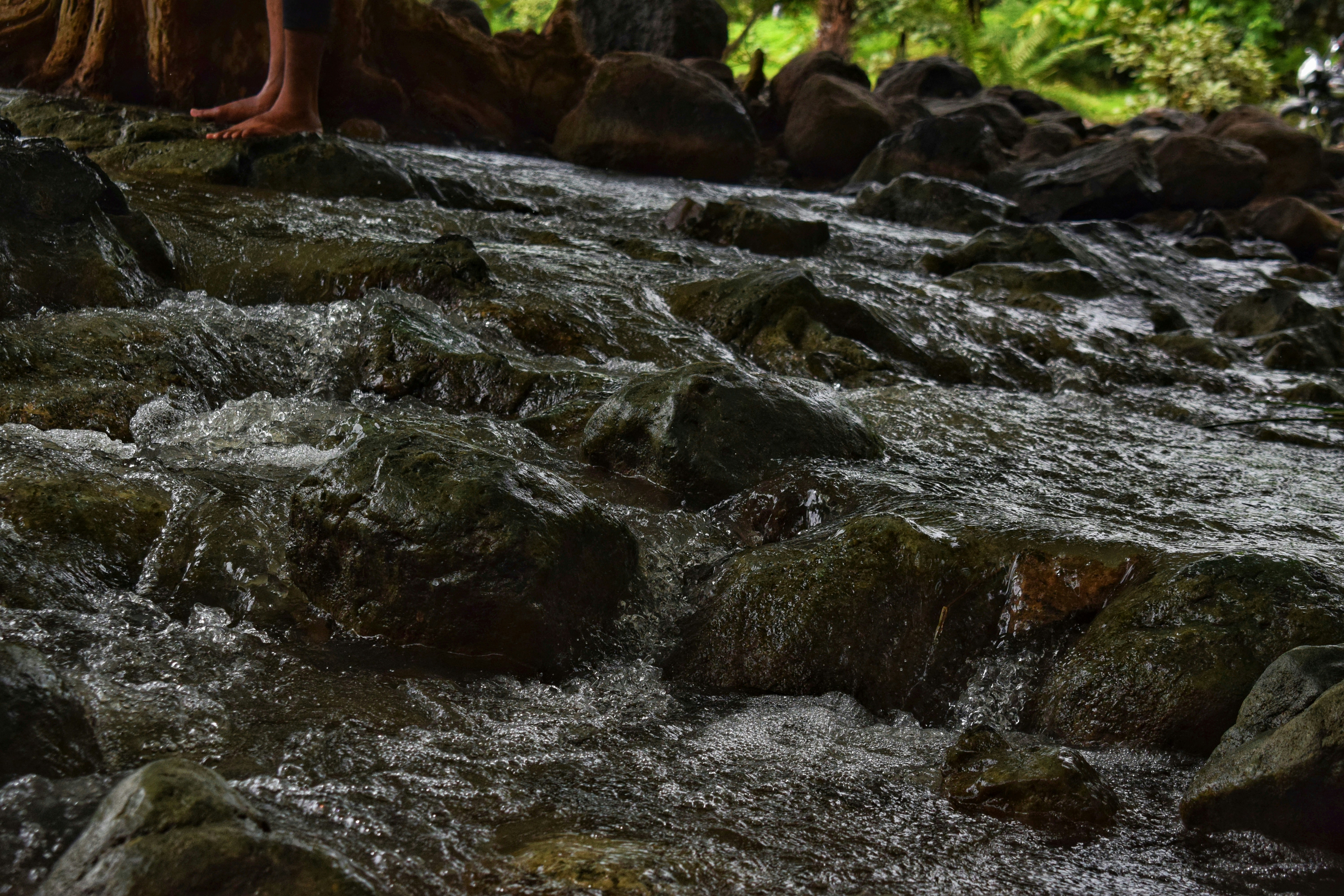 Clear water cascades over smooth stones, with a hint of a person's foot visible, illustrating the serene interaction between nature and humanity.