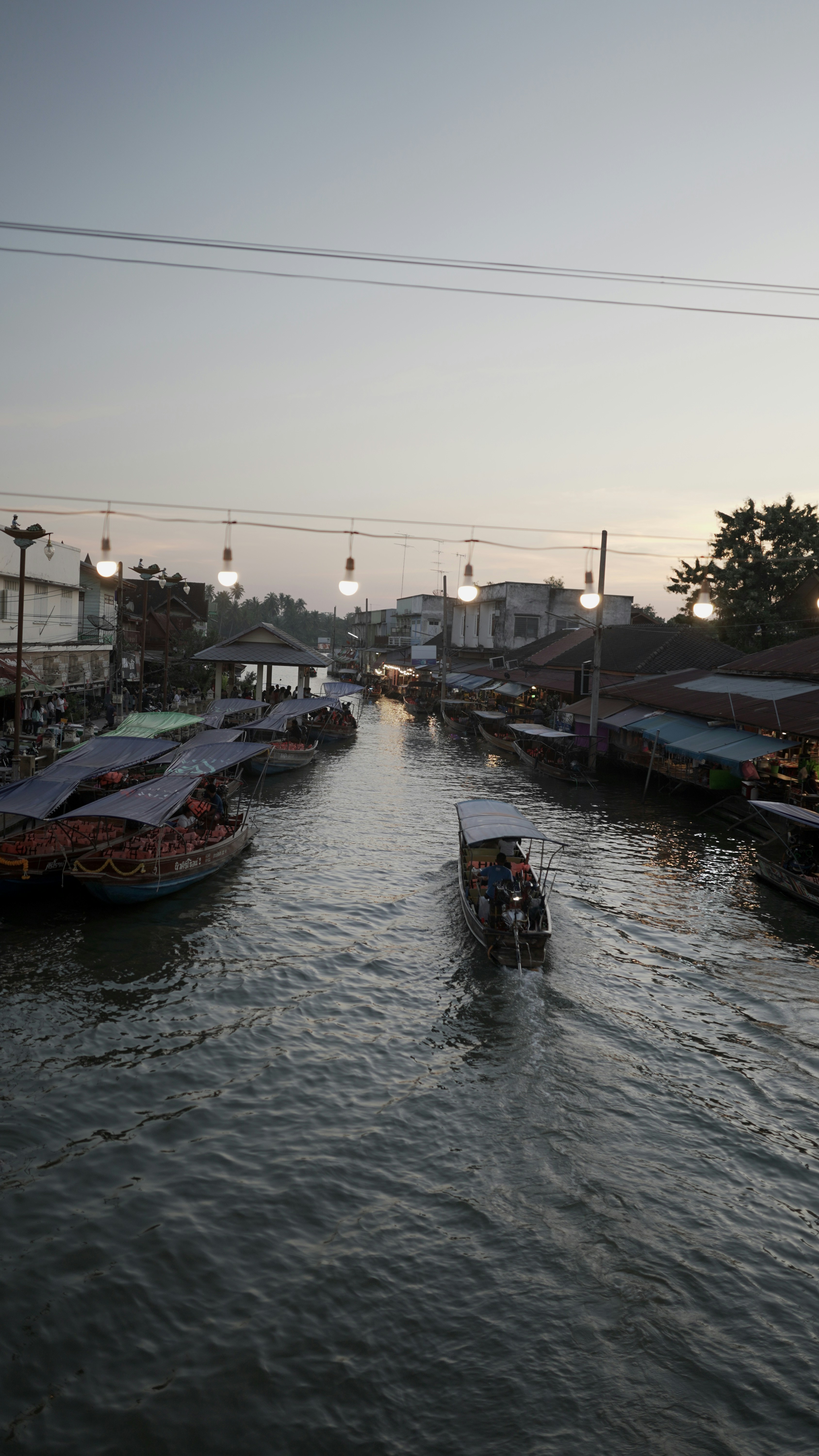Boats glide through a tranquil canal at dusk, with hanging lights illuminating the scene.