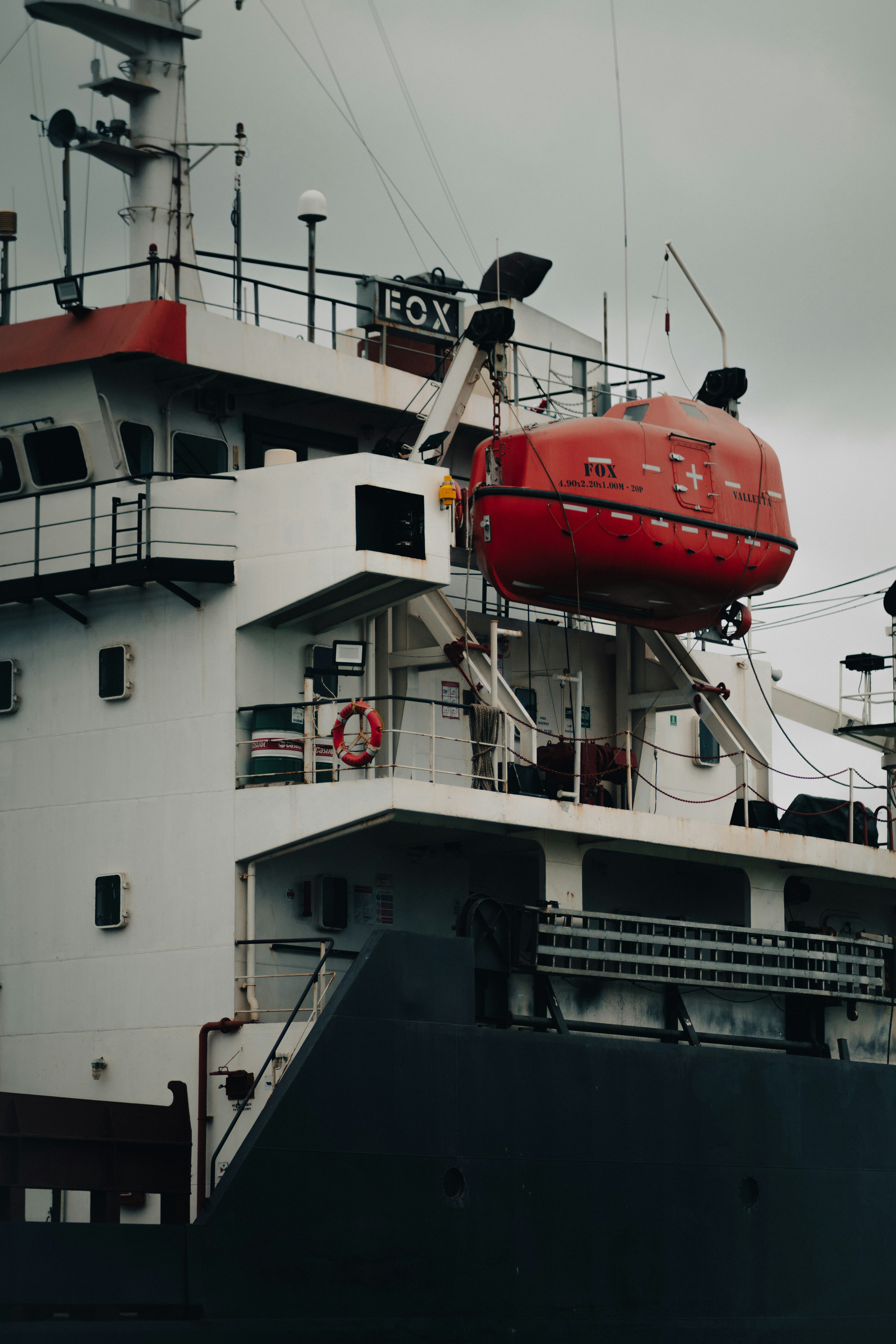 A cargo ship's deck featuring a bright orange lifeboat and safety equipment against a moody sky.