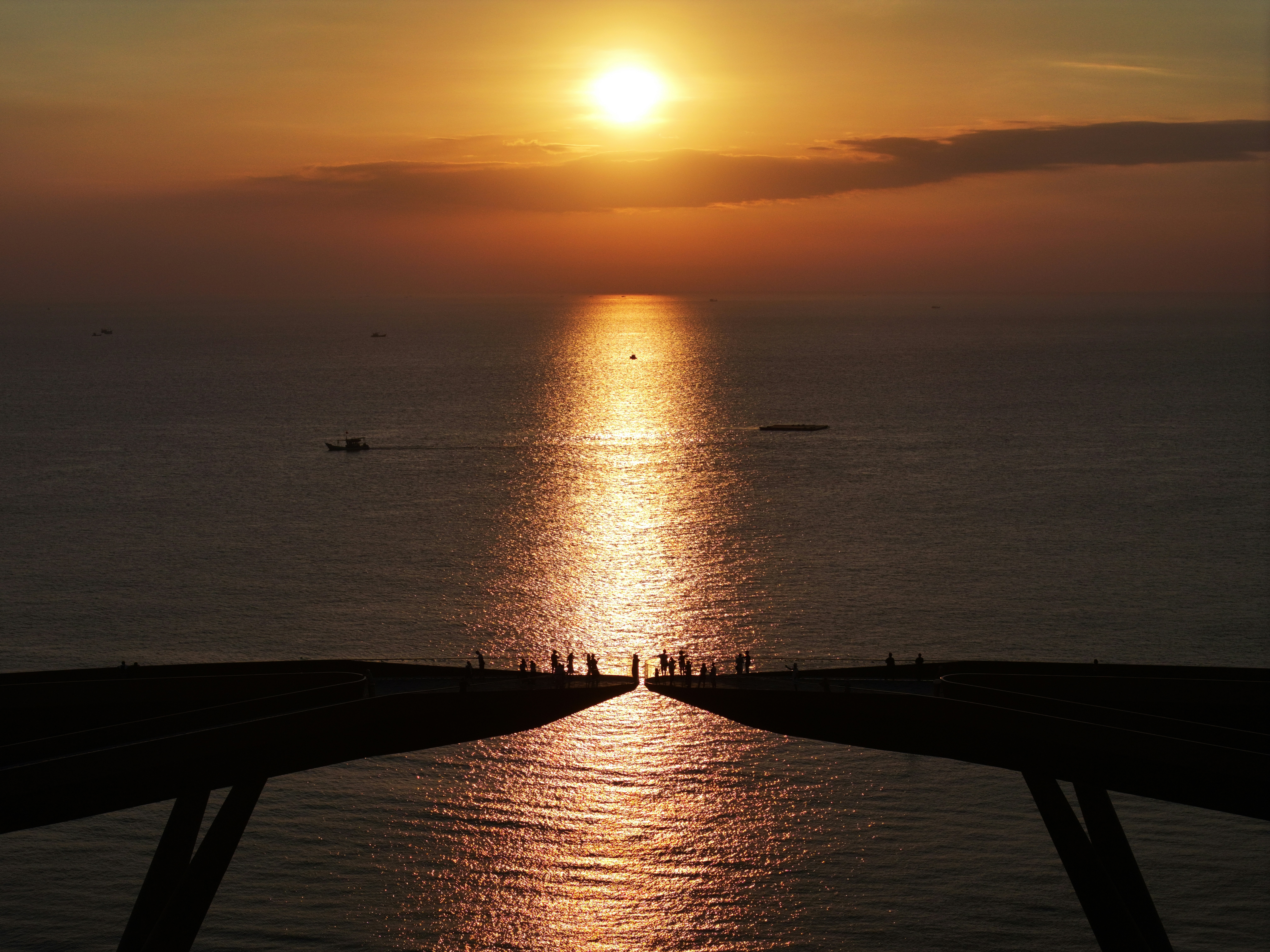 Sunset over a calm sea with silhouetted figures on a pier, a bright golden reflection stretching toward the foreground.