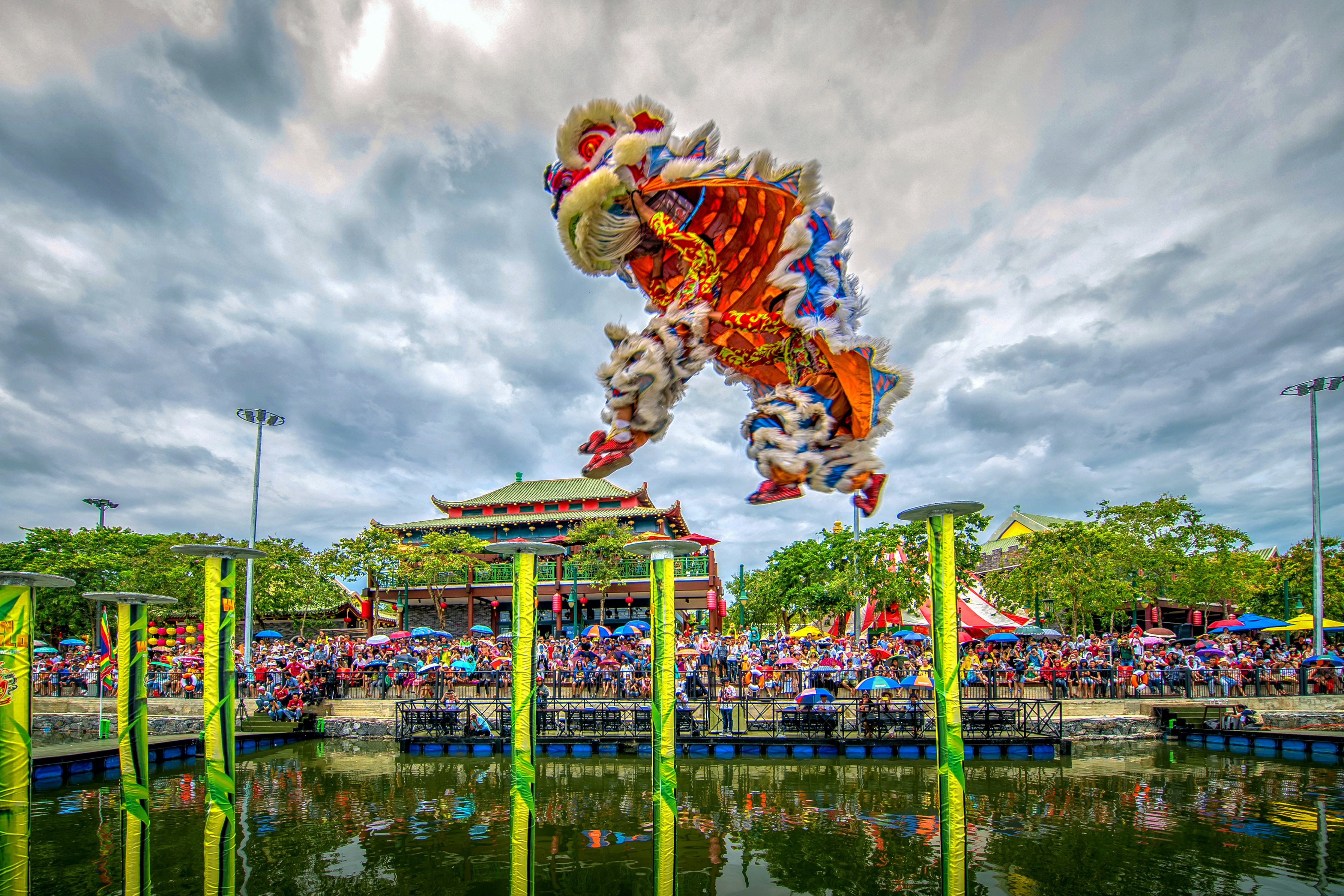 Colorful lion dance figure leaps dramatically over a reflective pond with a festive crowd in the background.