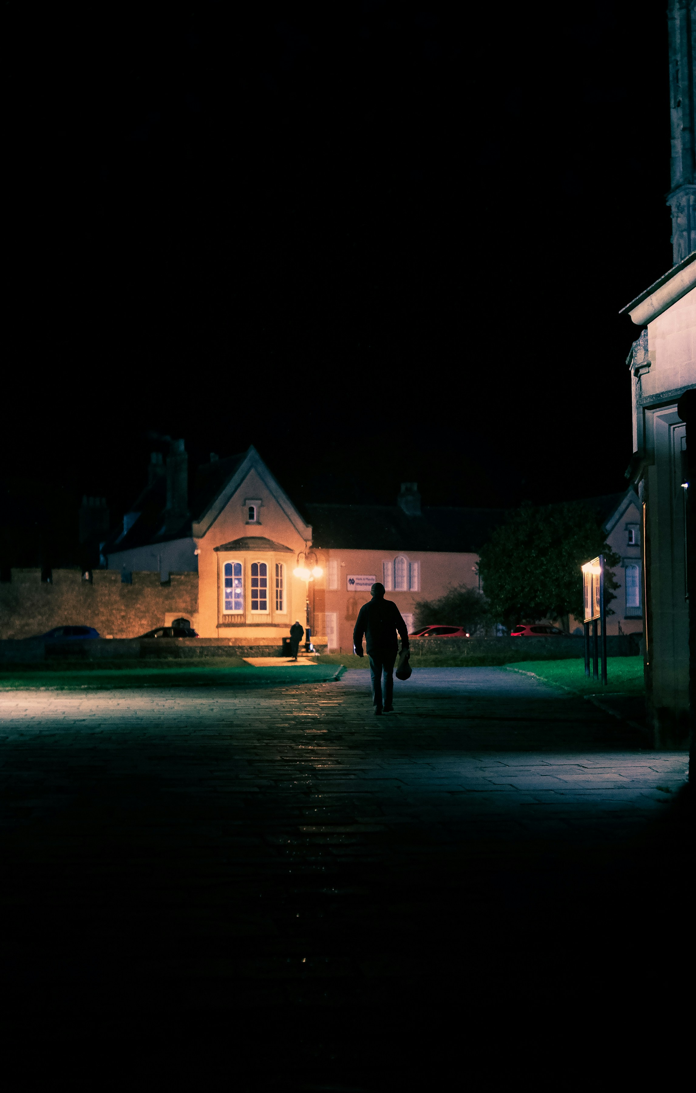 Silhouette of a person walking down a cobblestone path illuminated by warm streetlights, with charming buildings in the background. Nighttime ambiance enhances the scene.
