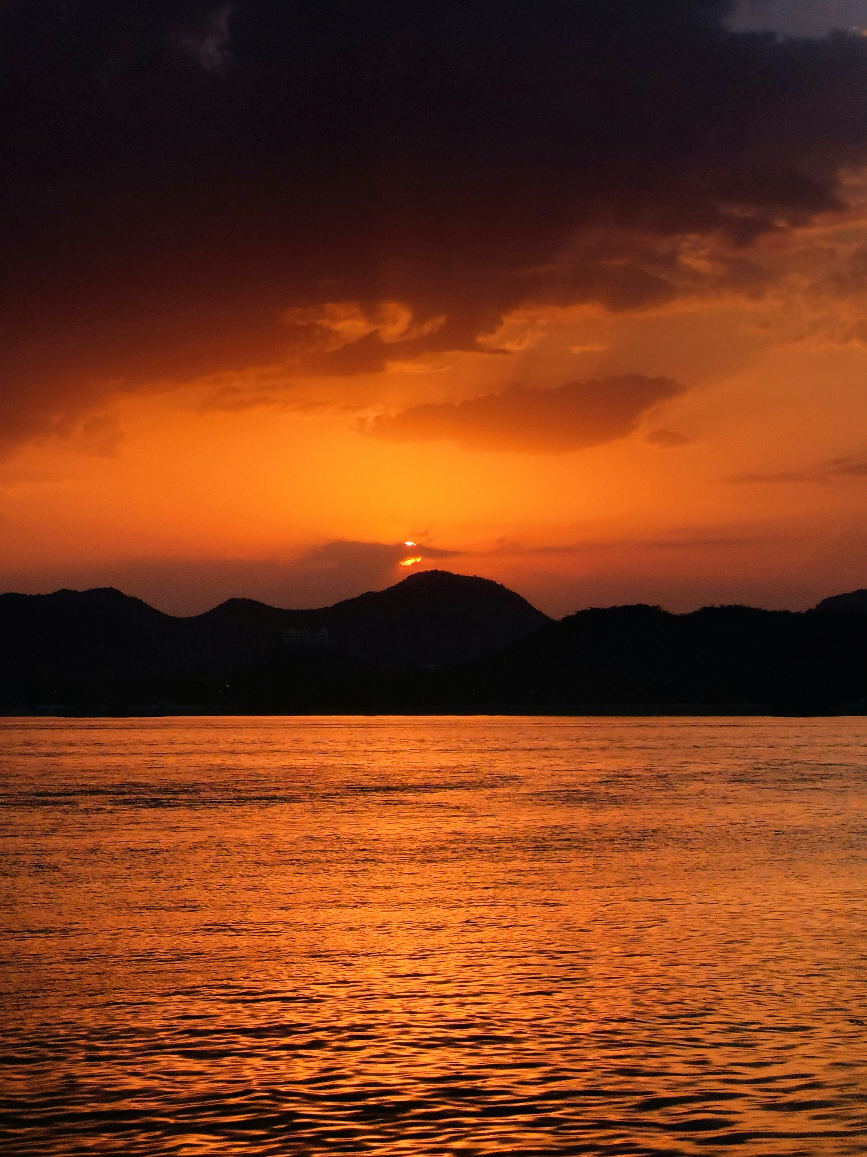 Golden sun setting behind mountains, casting warm reflections on the water's surface. The sky transitions from orange to deep purple, enhancing the tranquil atmosphere.