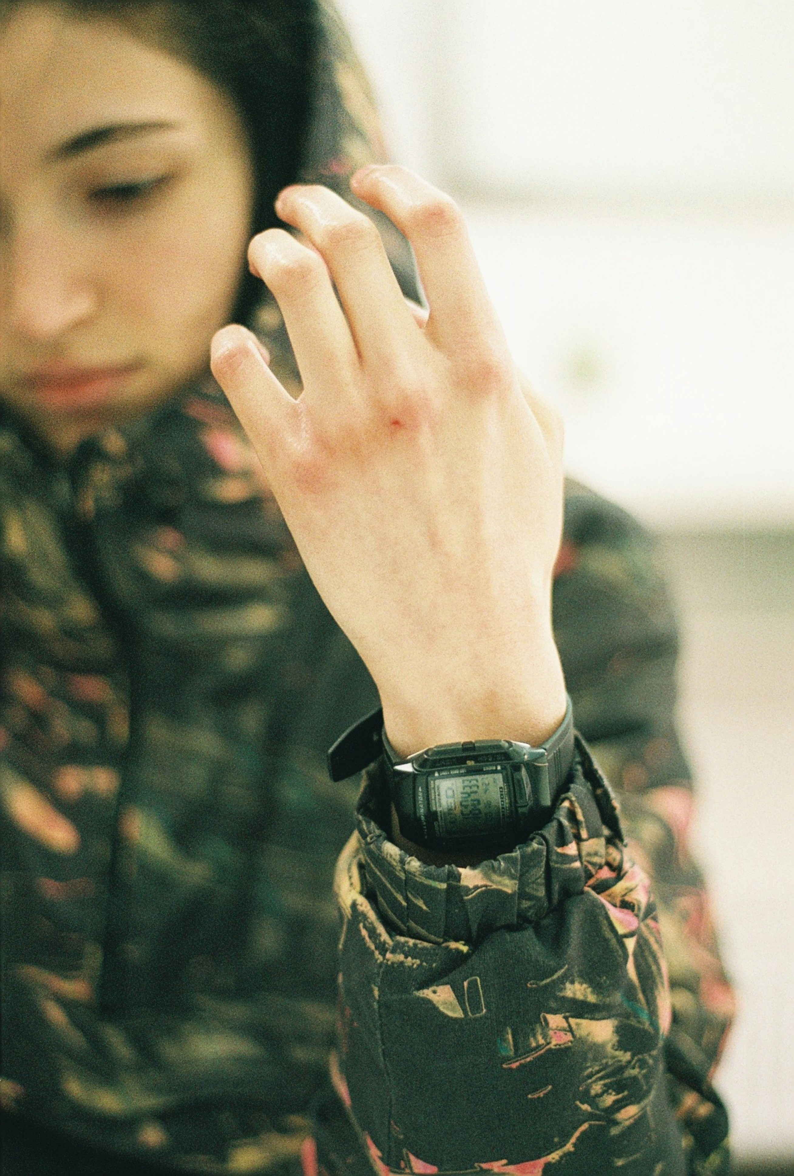 Close-up photograph of a hand adjusting a wristwatch on a camo jacket, with a softly blurred face in the background.