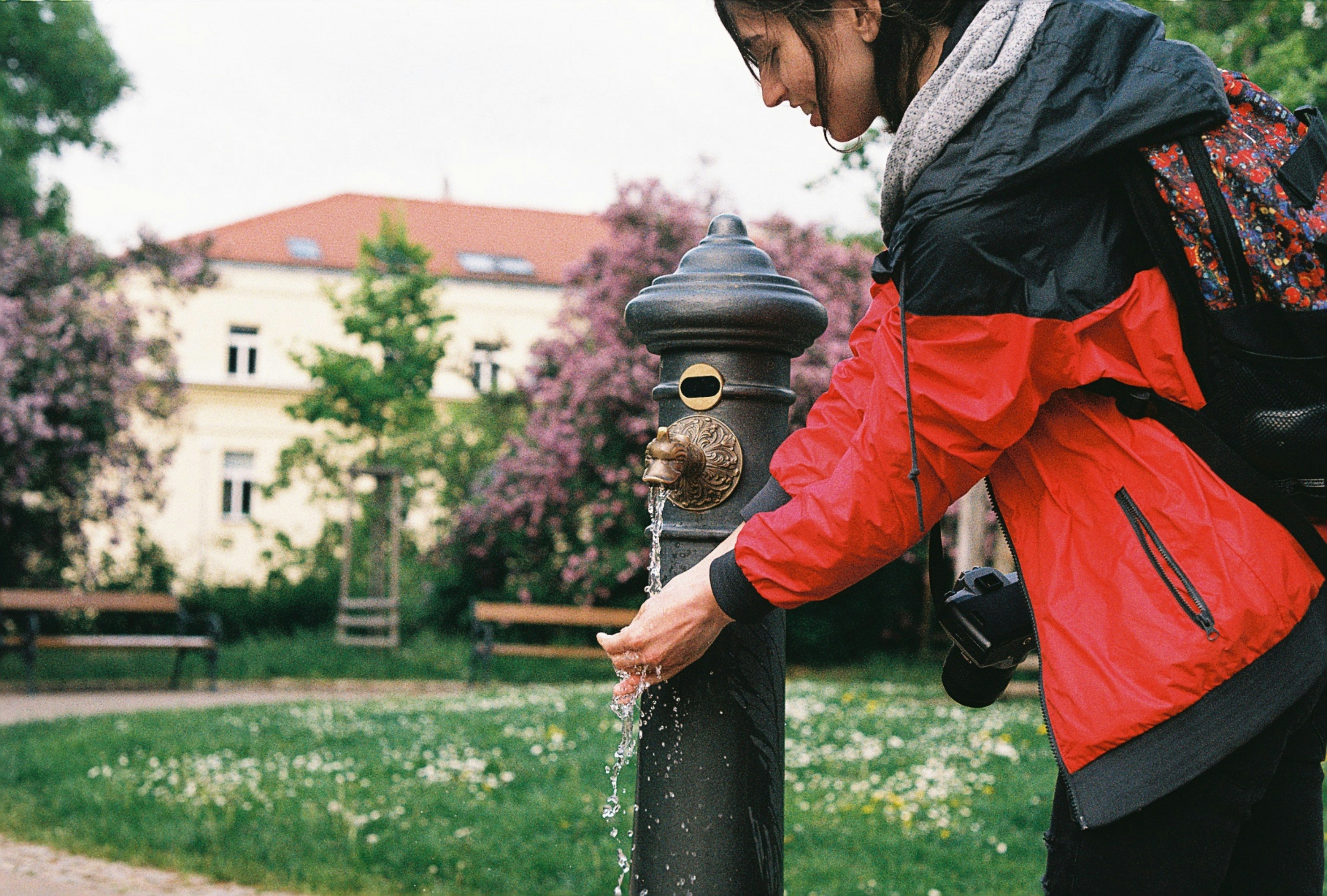 A girl washes her hands under a public faucet that is designed so beautifully. She is wearing a black and white Nike jacket. It is located in a park in Prague.