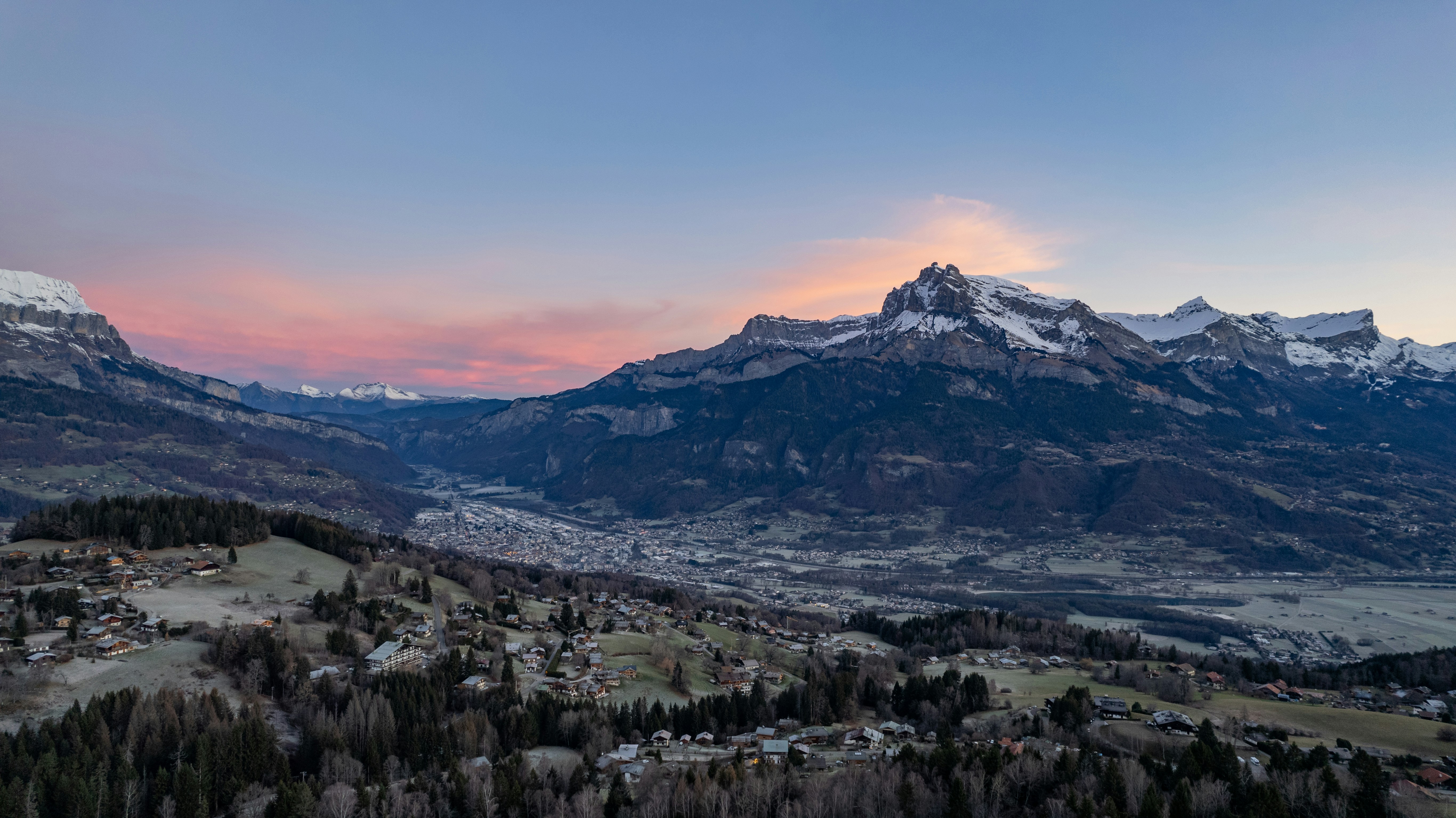 Snow-covered peaks of Mont Blanc massif under a pink sunrise sky, framed by alpine wilderness and evergreen trees.