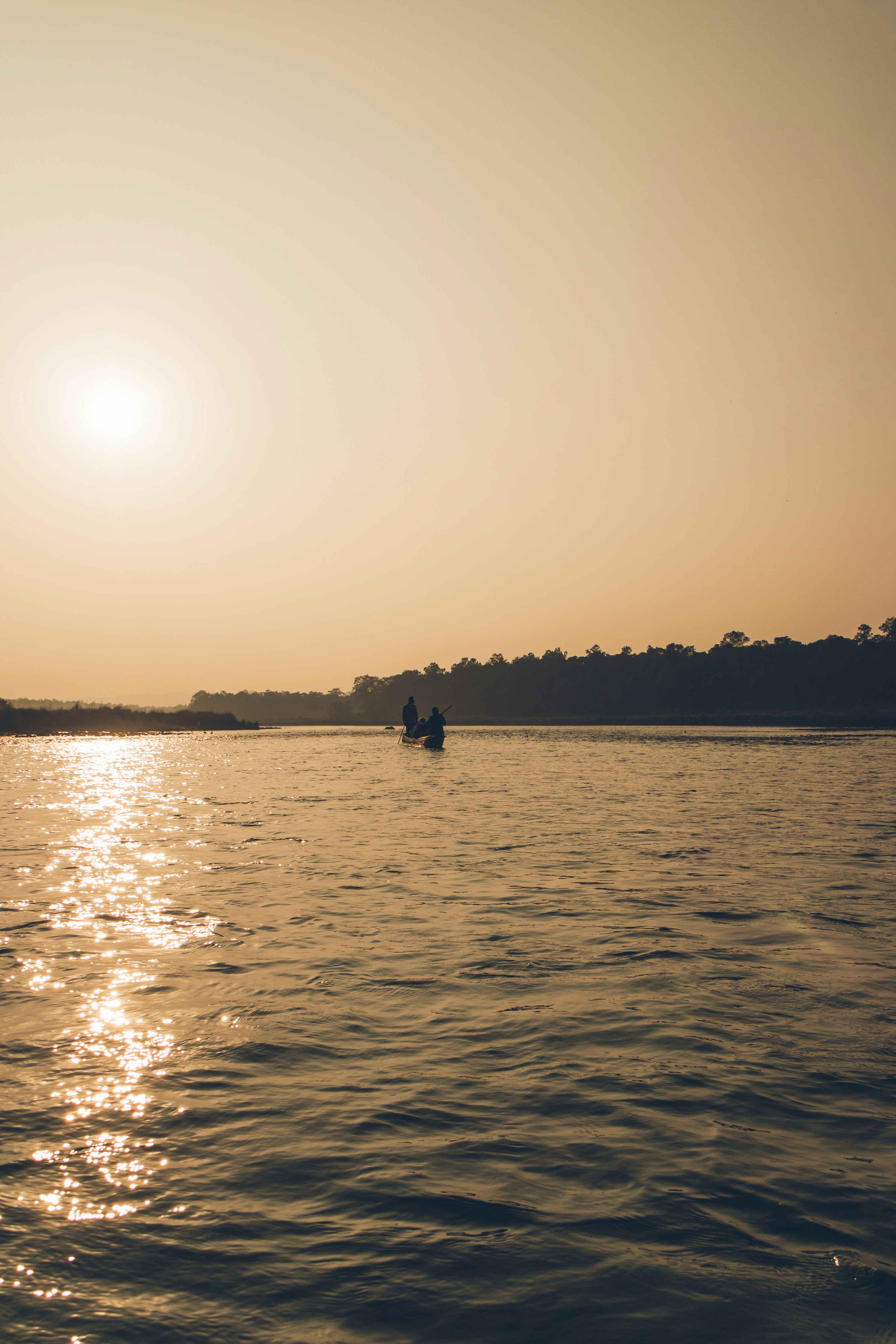Sunset over a calm river with a small canoe carrying two people, reflections of golden light ripple across the water and a tree-lined shore on the horizon.