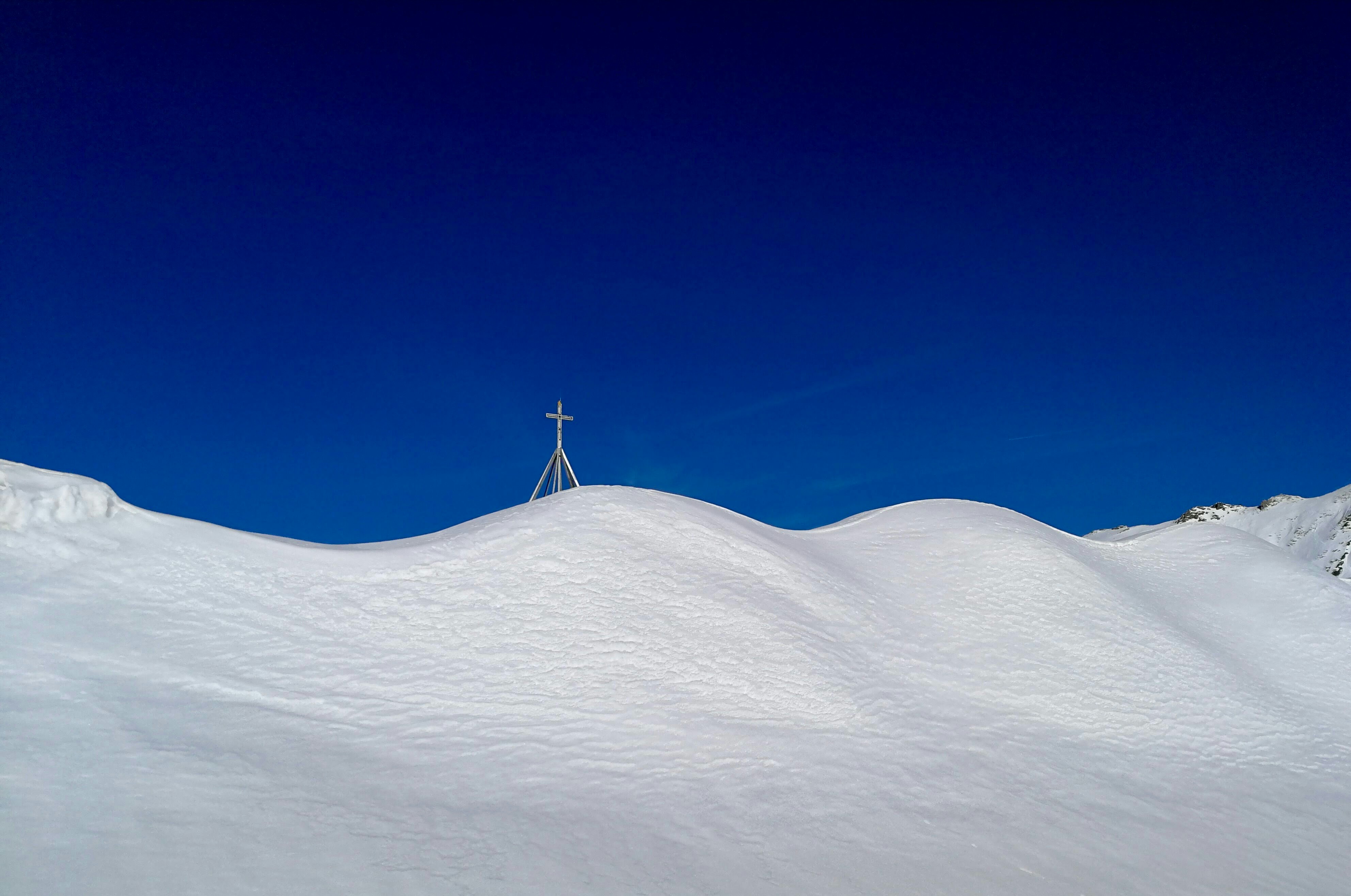 A lone cross sits atop a gentle snow ridge as a deep blue gradient sky stretches overhead.