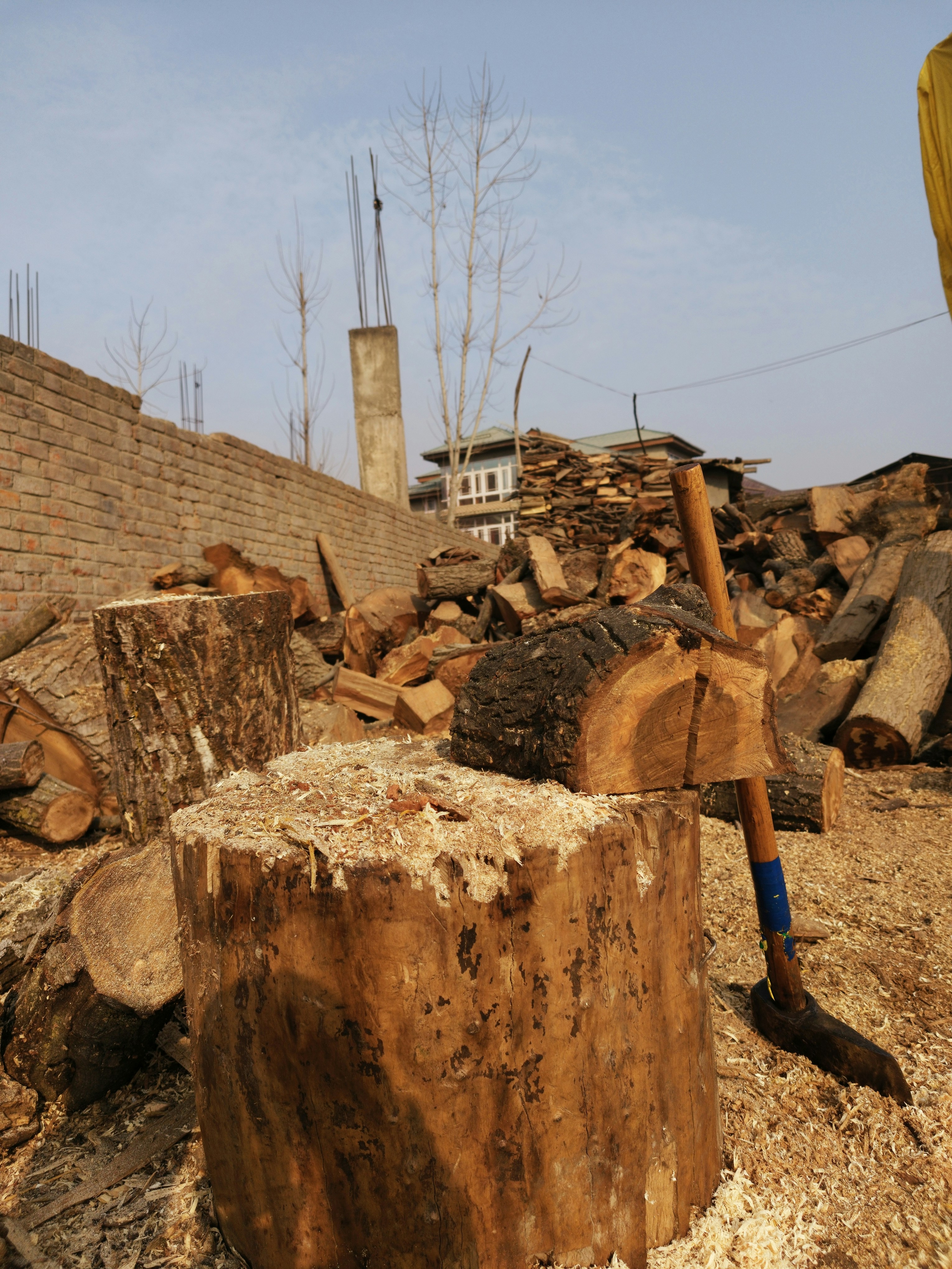 Photo: A blue-taped axe rests in a pile of chopped logs and a large stump, set against a brick wall with scattered timber under a pale blue sky.