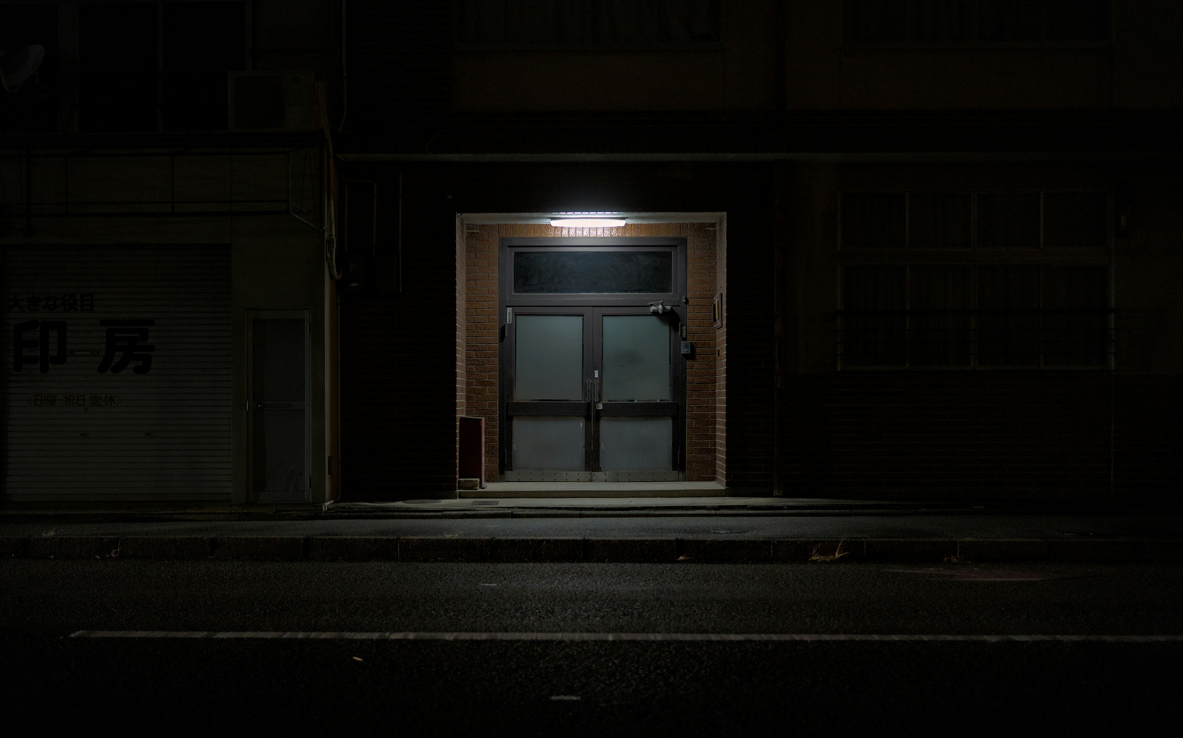 The lamp above a building entrance illuminates the pavement and street in front of it, while the rest of the building remains ominously dark. 2024-01-09 @ XPM8+PM3Julien