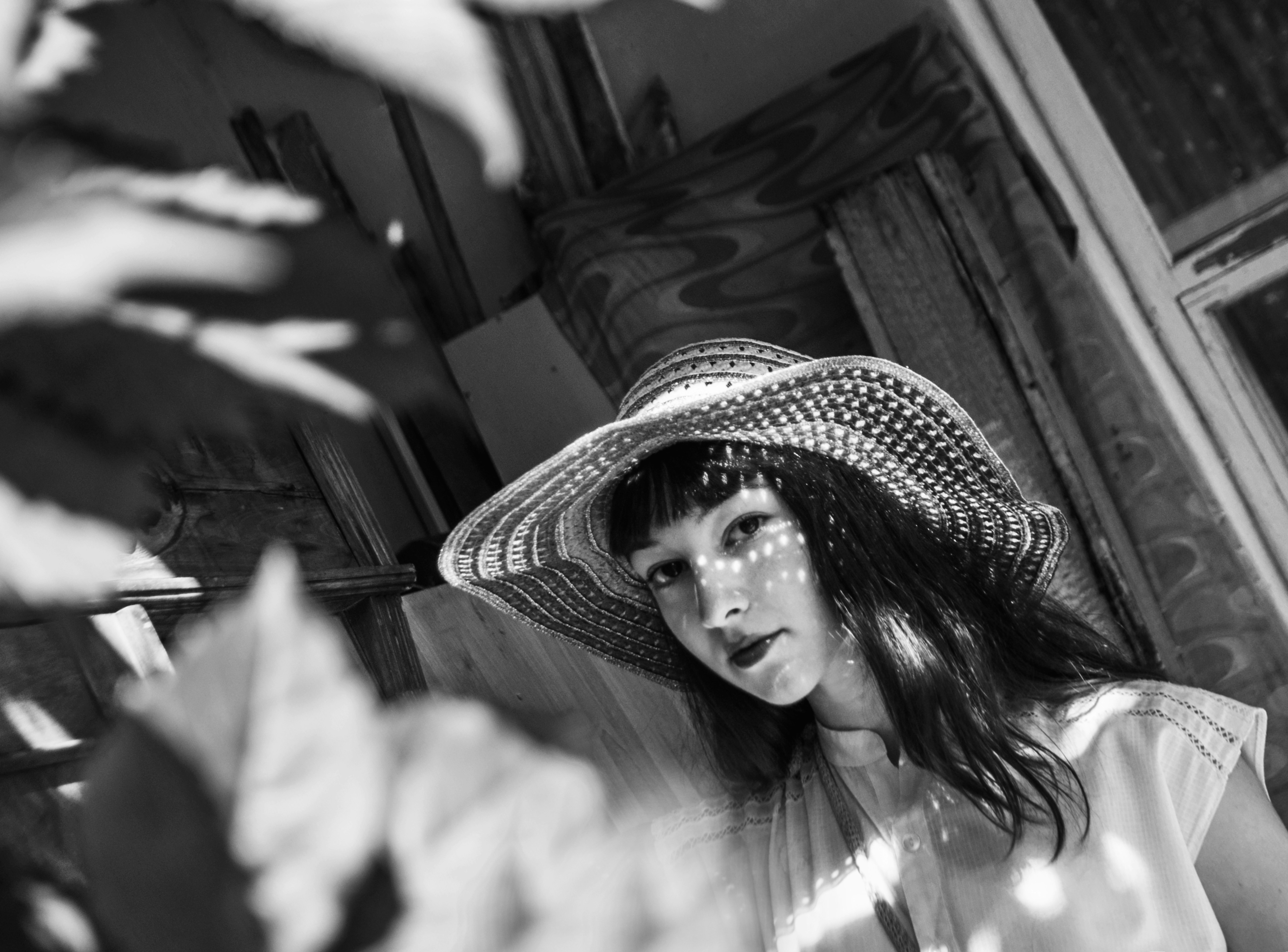 Woman wearing a wide-brimmed hat surrounded by soft-focus leaves in a black and white setting.