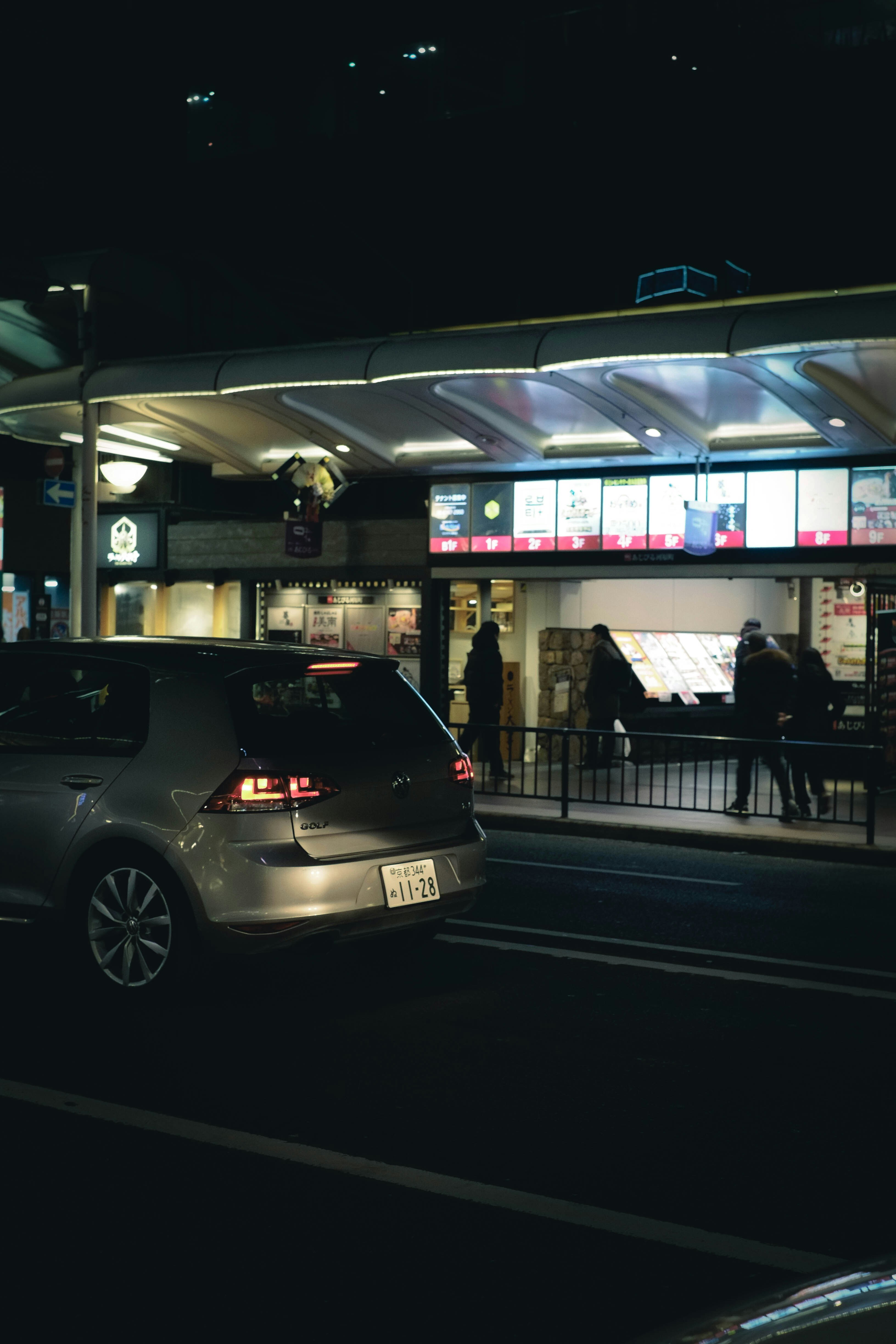 Midnight curbside: Silver hatchback beside a glowing fast-food kiosk under an illuminated canopy