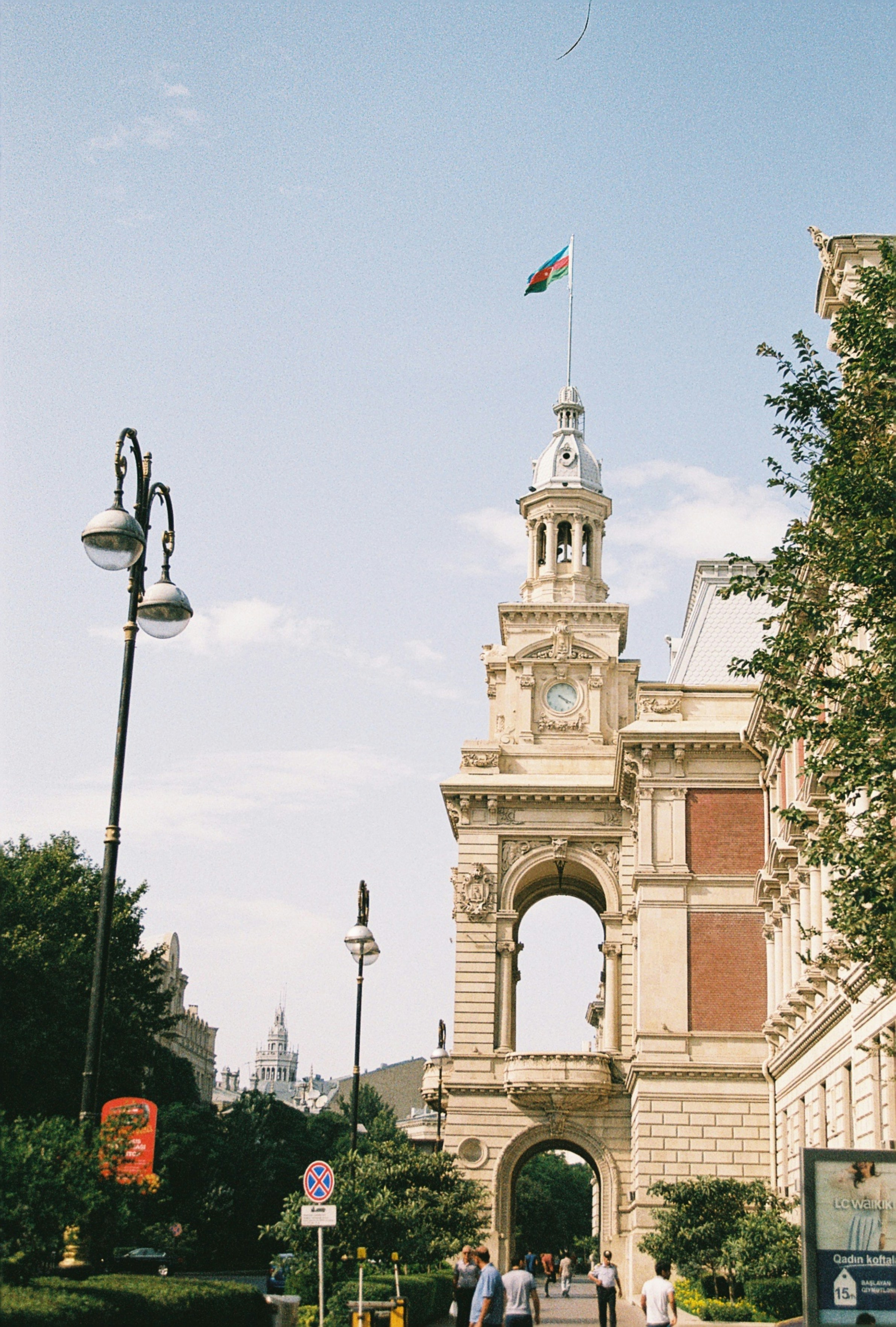 Daylight photograph of a grand archway gate dominating a city street, with a flag fluttering atop the tower. Pedestrians stroll beneath lampposts while trees frame the scene against a clear sky.