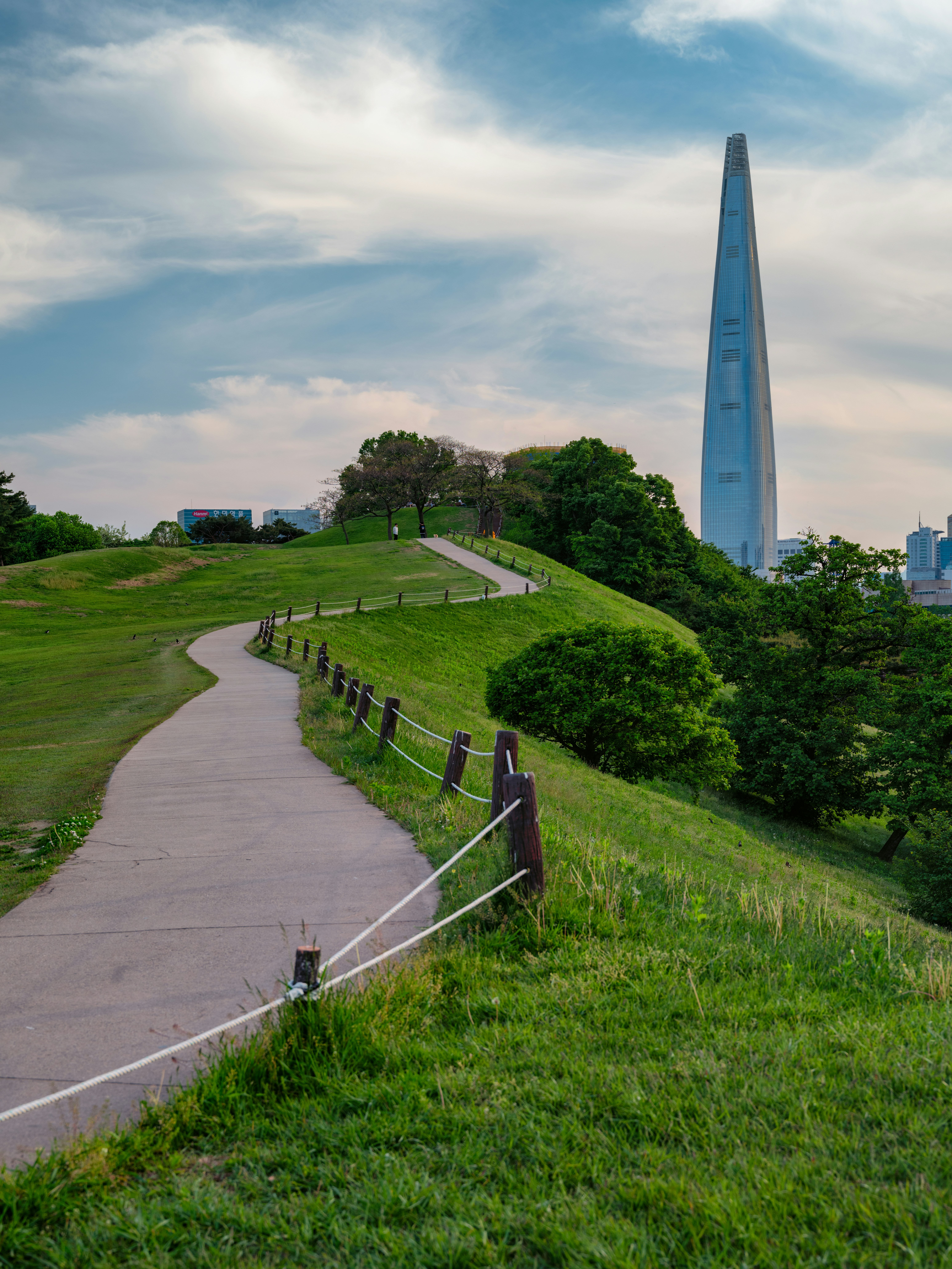 A meandering paved path climbs a grassy hill toward a distant glass skyscraper, framed by trees under a bright blue sky.