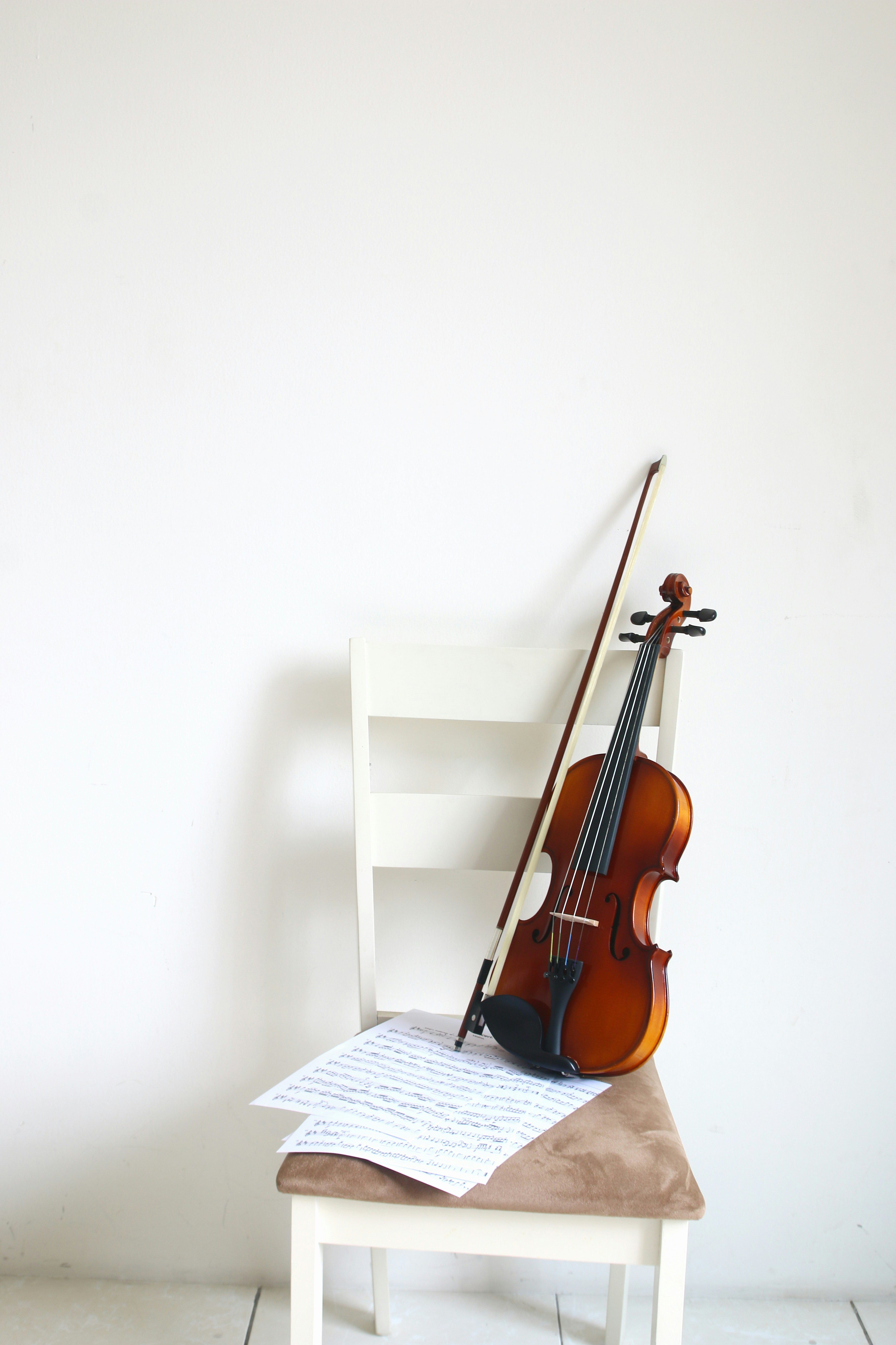 A violin rests gracefully on a beige chair, accompanied by sheets of music, set against a minimalistic white background.