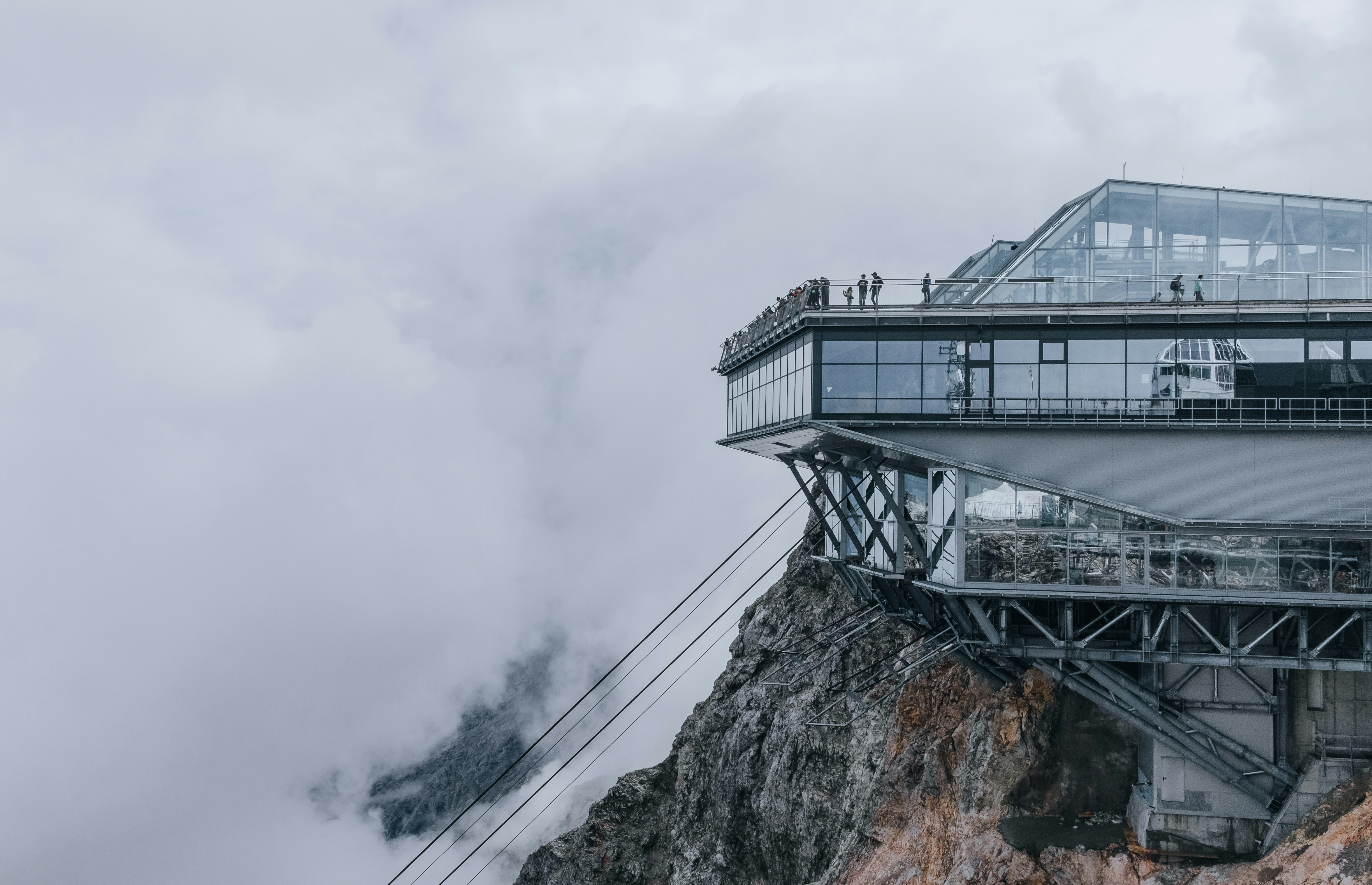 People stand at a glass viewing platform jutting from a rocky cliff, with fog wrapping the scene and cables extending down the precipice.