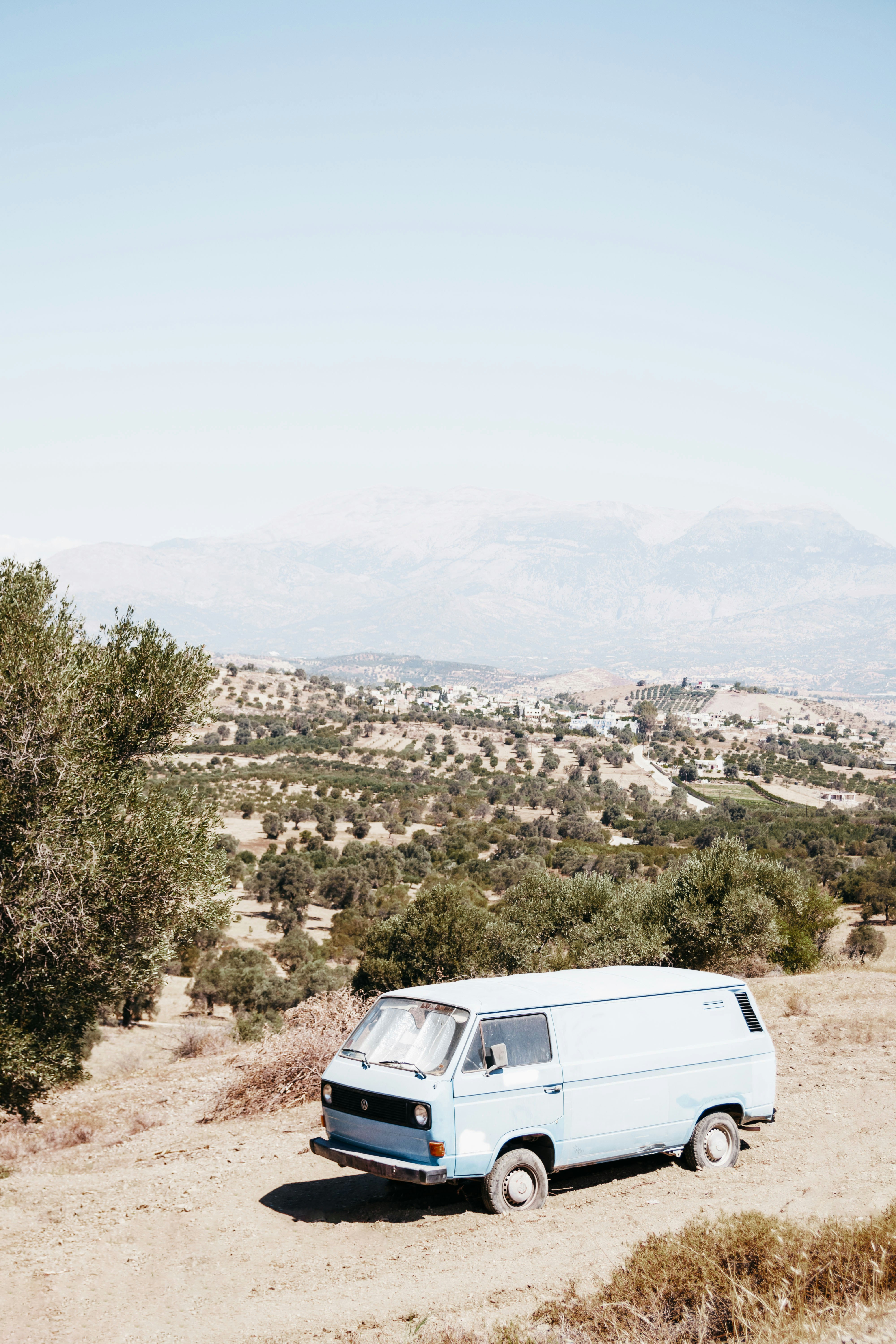 A vintage blue van parked on a hillside, surrounded by olive trees and rolling landscapes under a clear sky.