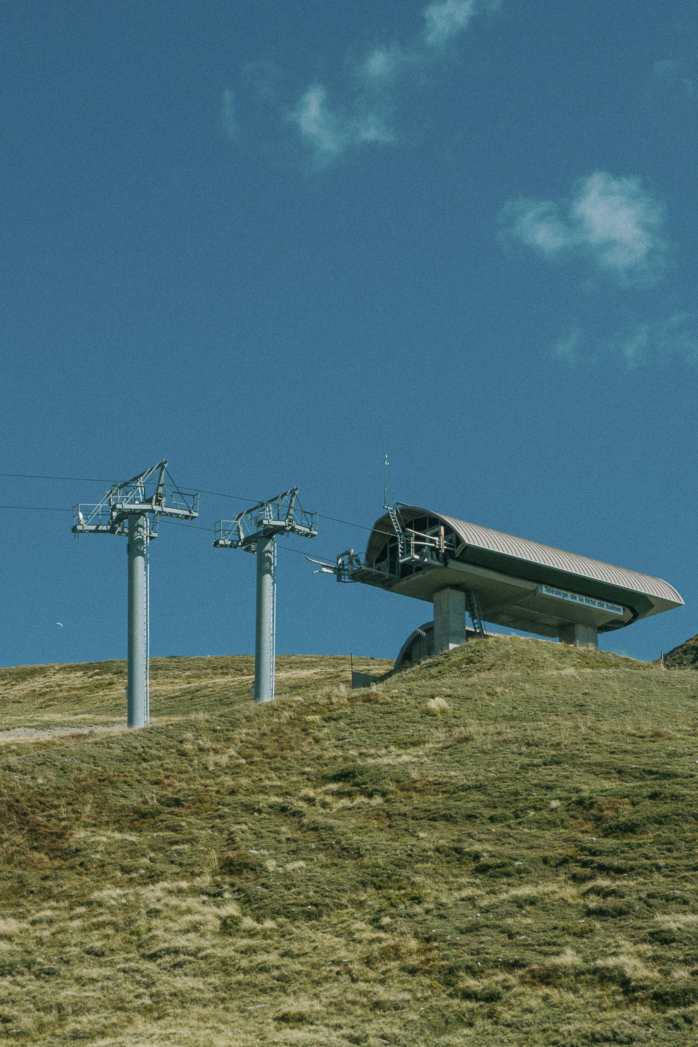 Hilltop ski-lift station with two supporting towers rises over a grassy slope, set against a clear blue sky.