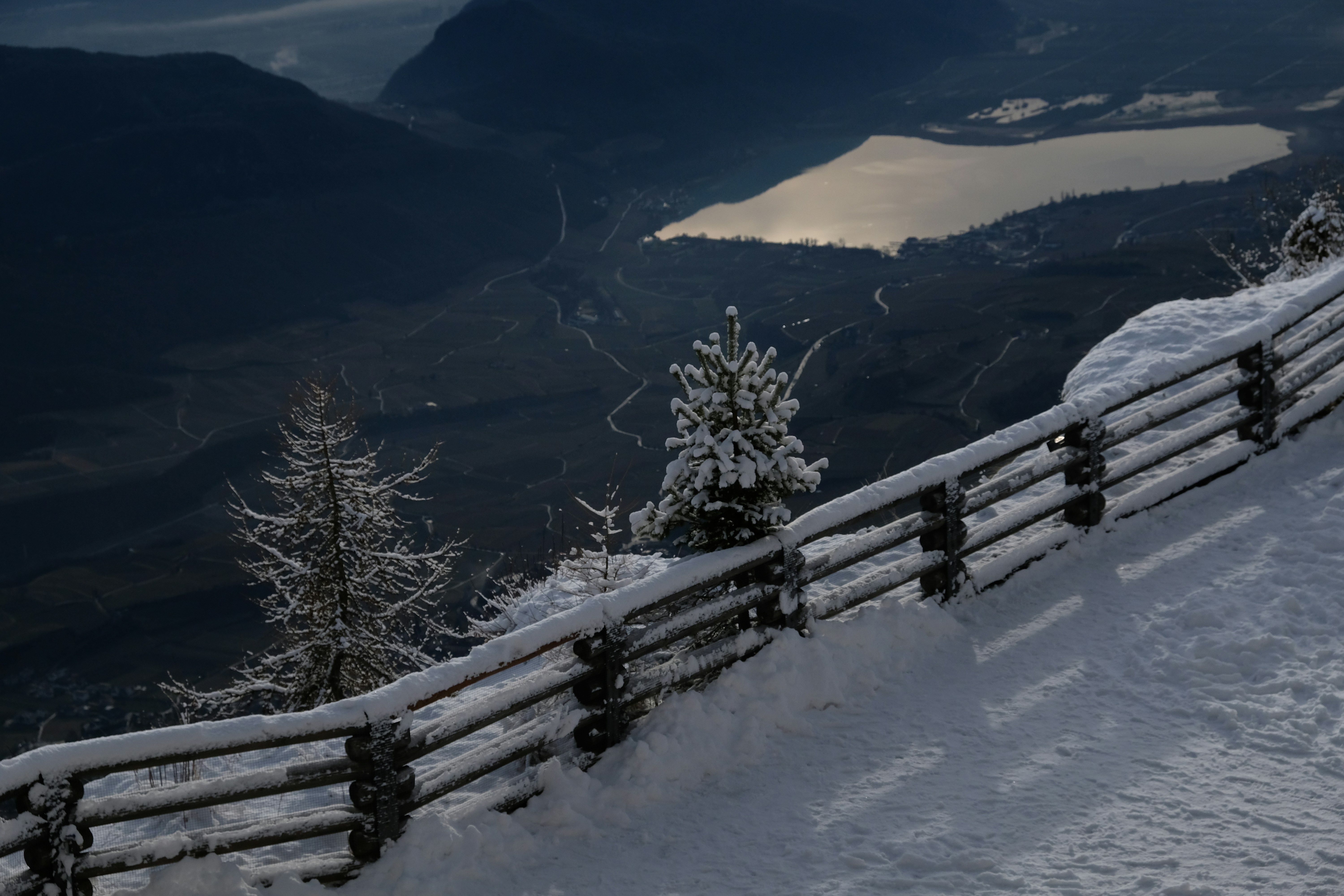 Snow-covered fence overlooking a serene mountain valley and distant lake under a darkened sky.