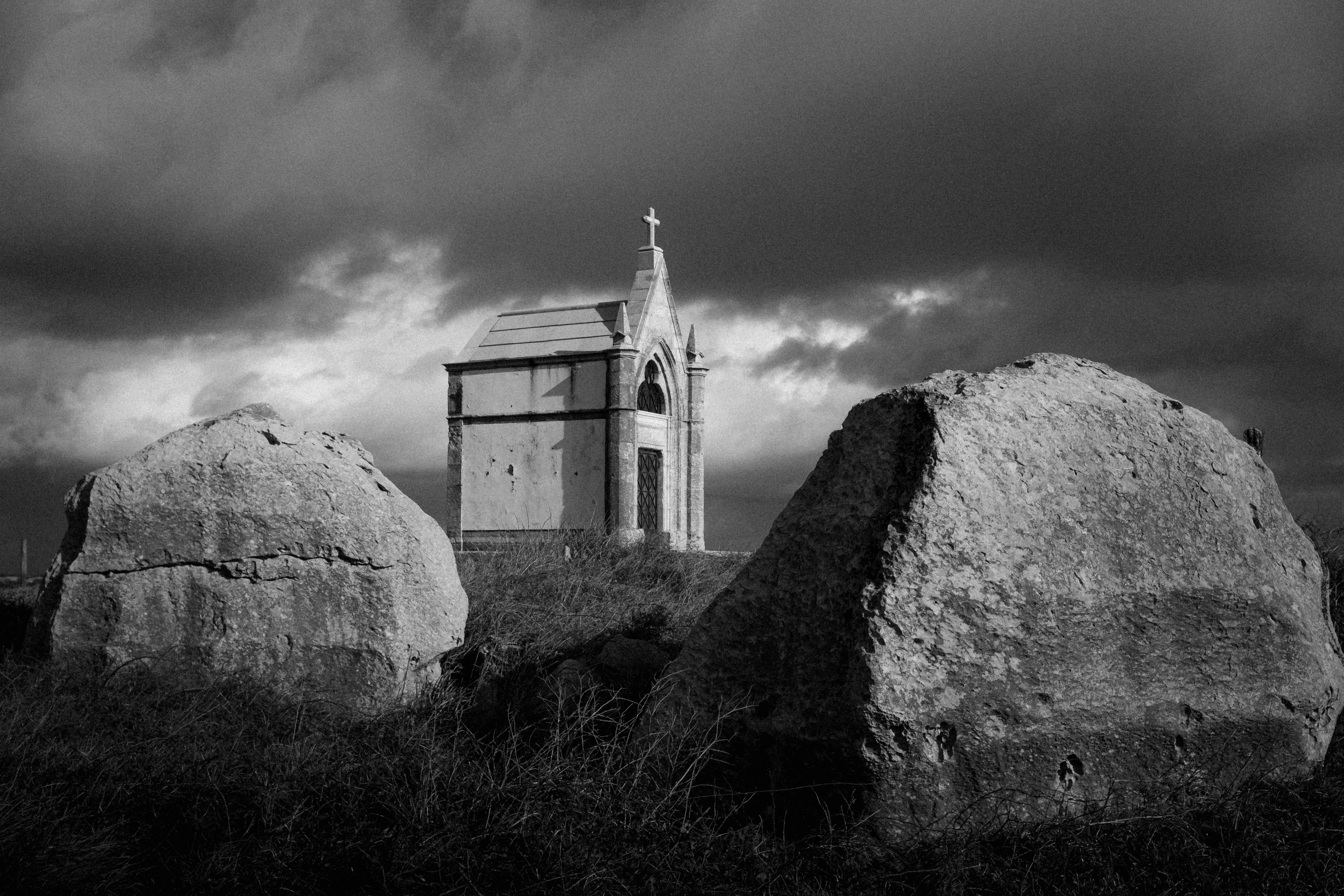 Ancient stone structure stands beneath dramatic, cloudy skies flanked by large boulders.