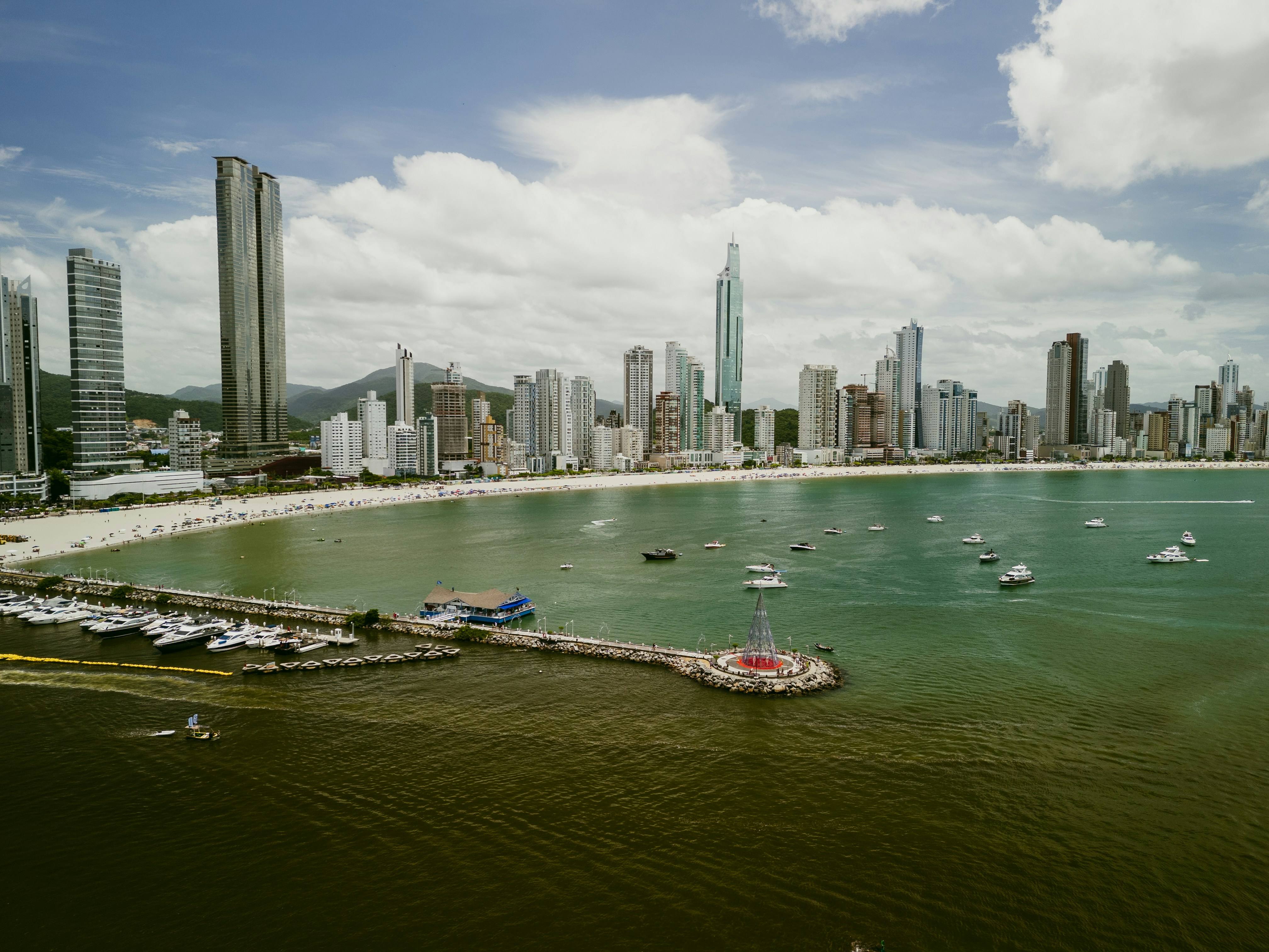 High-rise buildings lining a curving beach with boats dotting the turquoise waters under a partly cloudy sky.