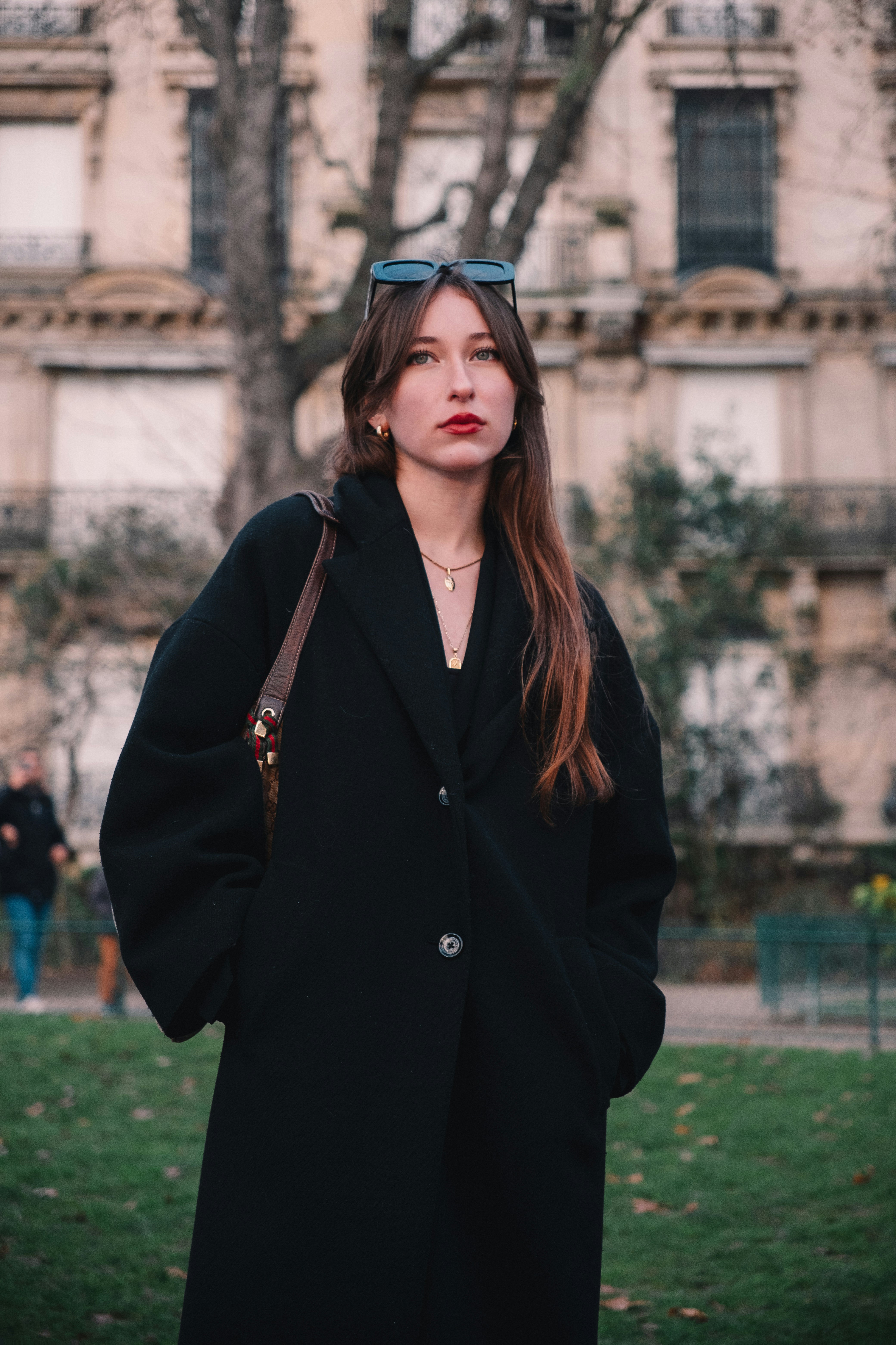 Portrait photograph of a woman in a long black coat standing in a park with historic façades behind; sunglasses rest on her head and she gazes forward.