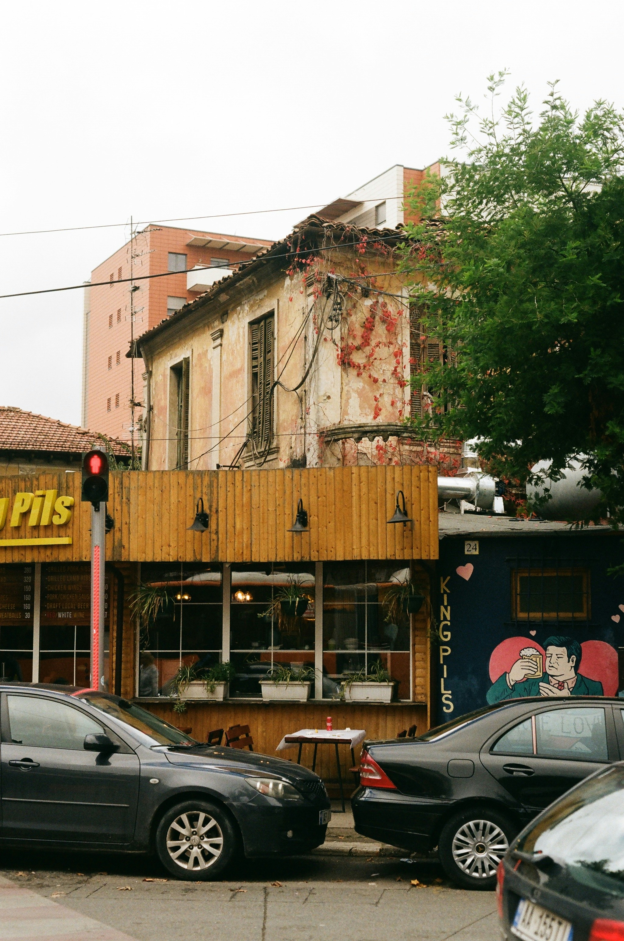 Ivy-clad corner café on a weathered brick block with a wooden storefront. A mural and parked cars line the street.