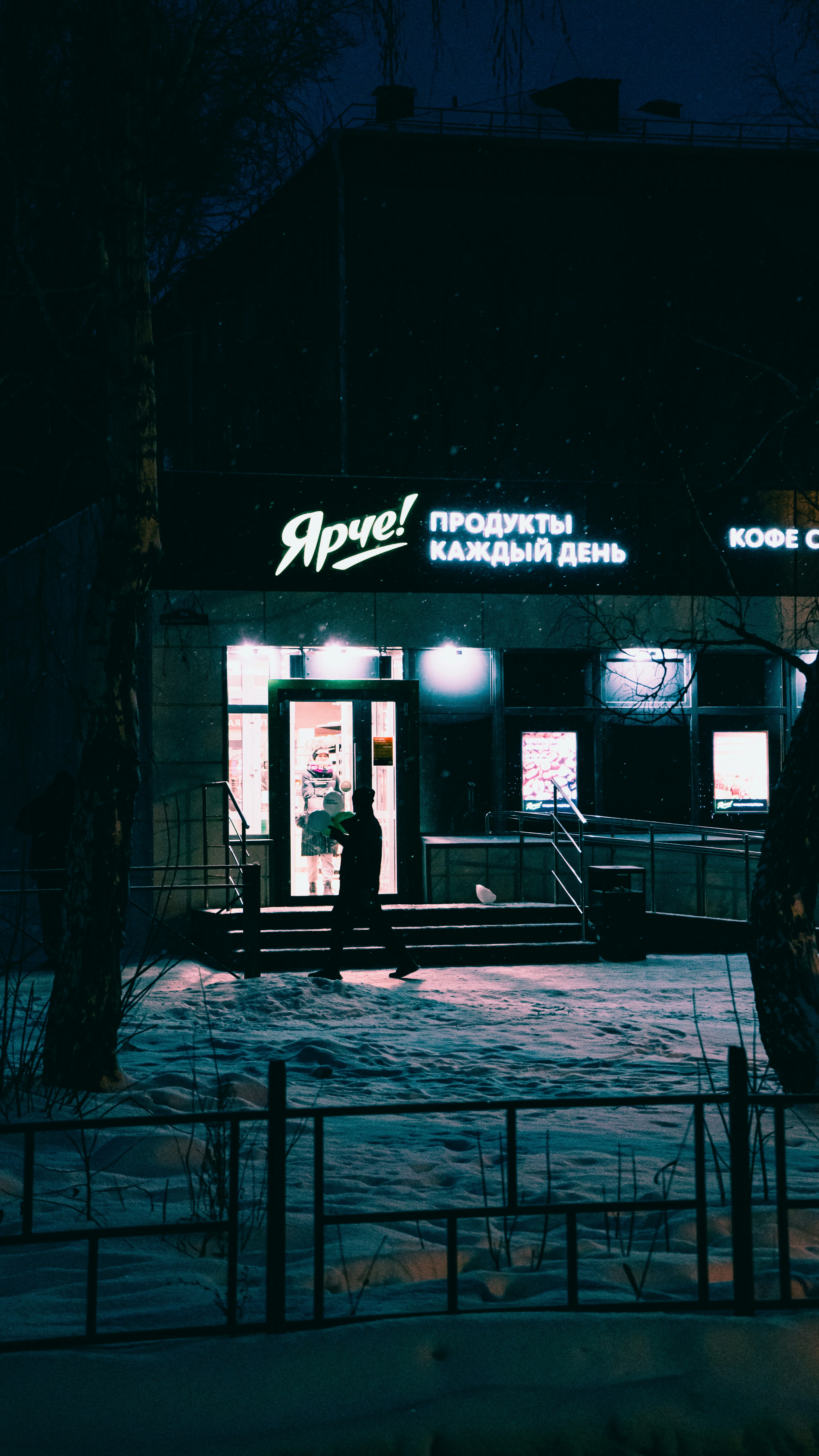 A person walks towards a brightly lit grocery store entrance amidst a snowy scene at night.