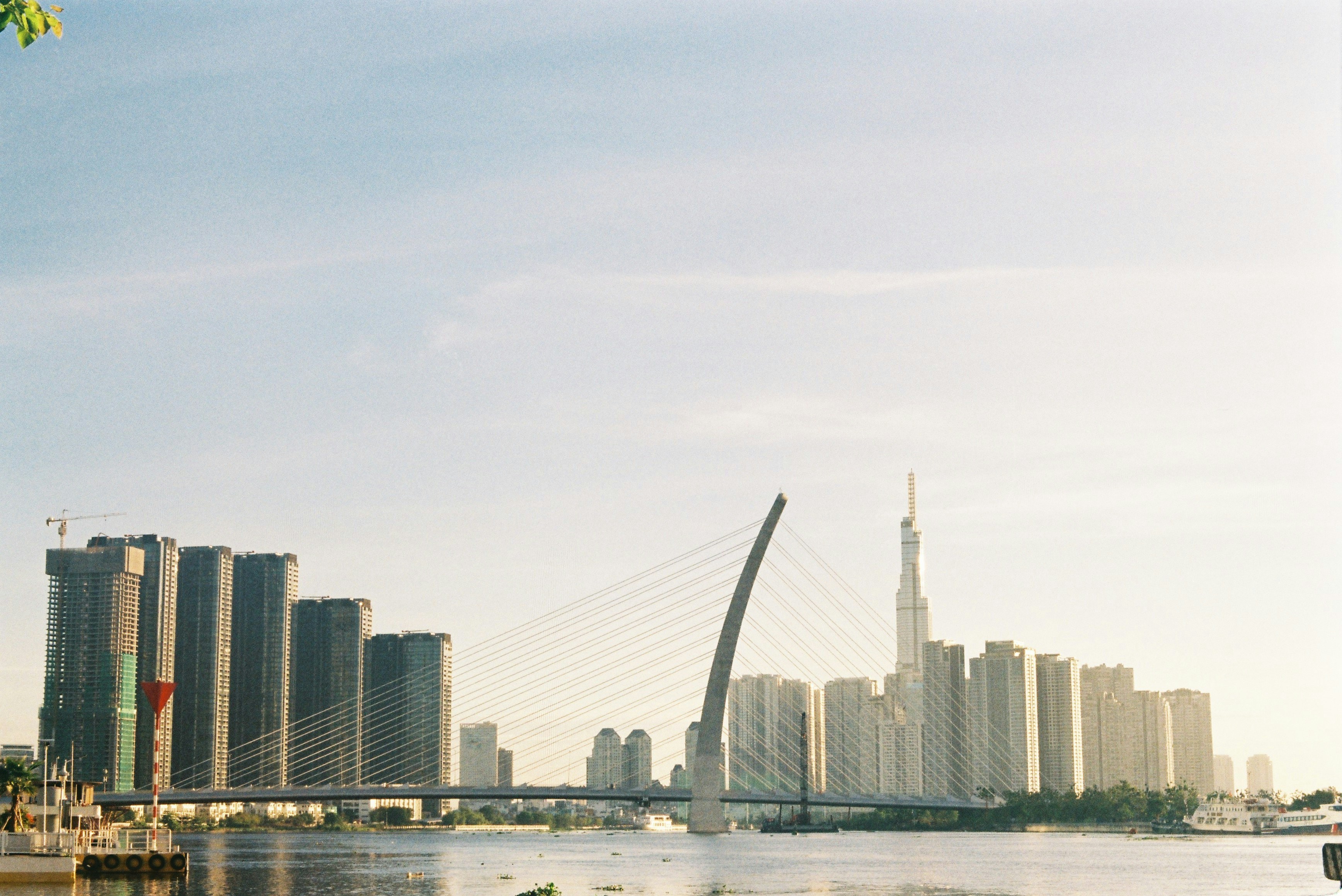 Modern suspension bridge arches gracefully over a serene river, framed by towering skyscrapers under a clear sky.