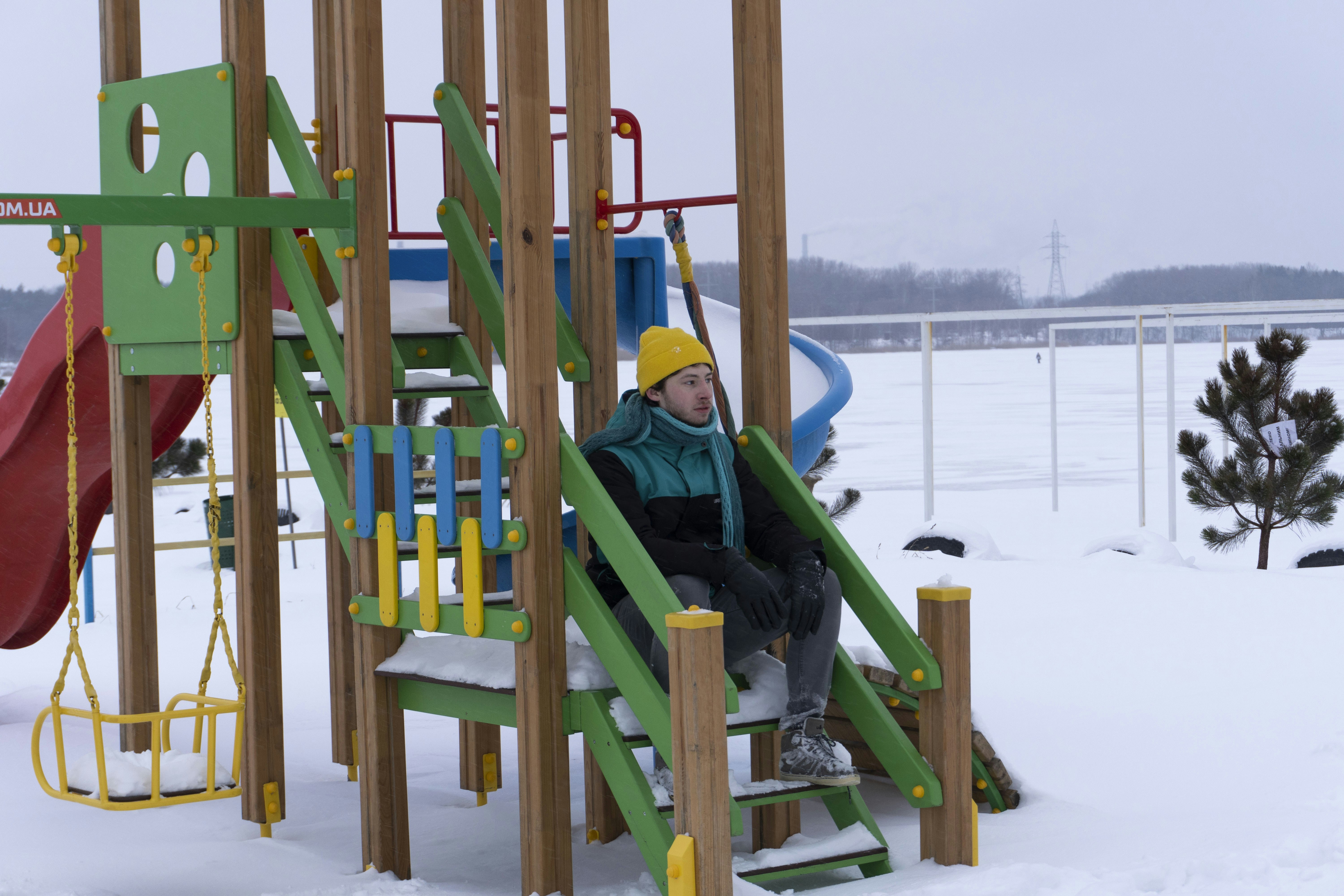 Winter Pause on a Green Climber: Child in Yellow Hat on Snowy Playground