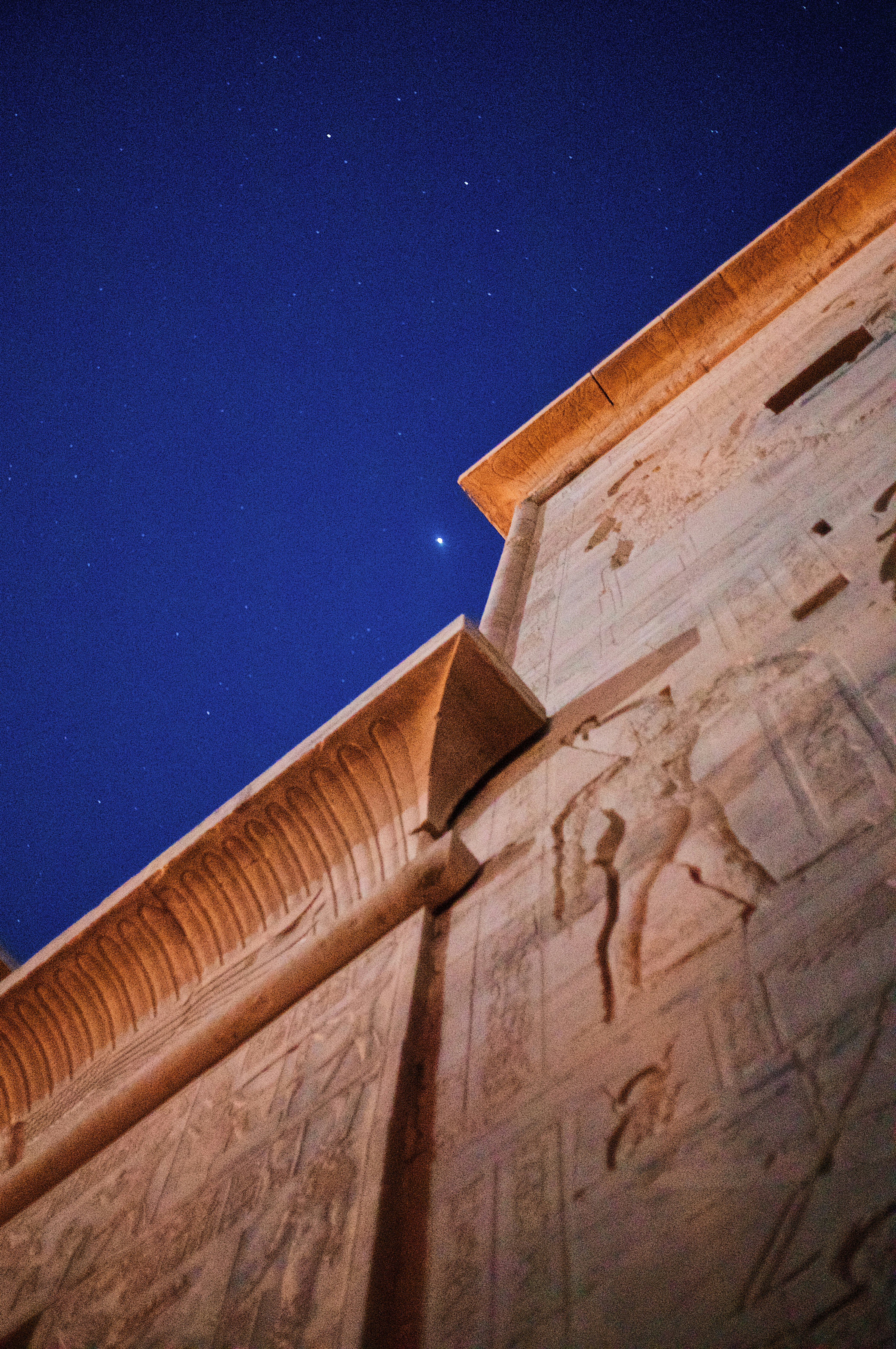 Intricate carvings on an ancient wall illuminated by soft light, set against a deep blue night sky dotted with stars.