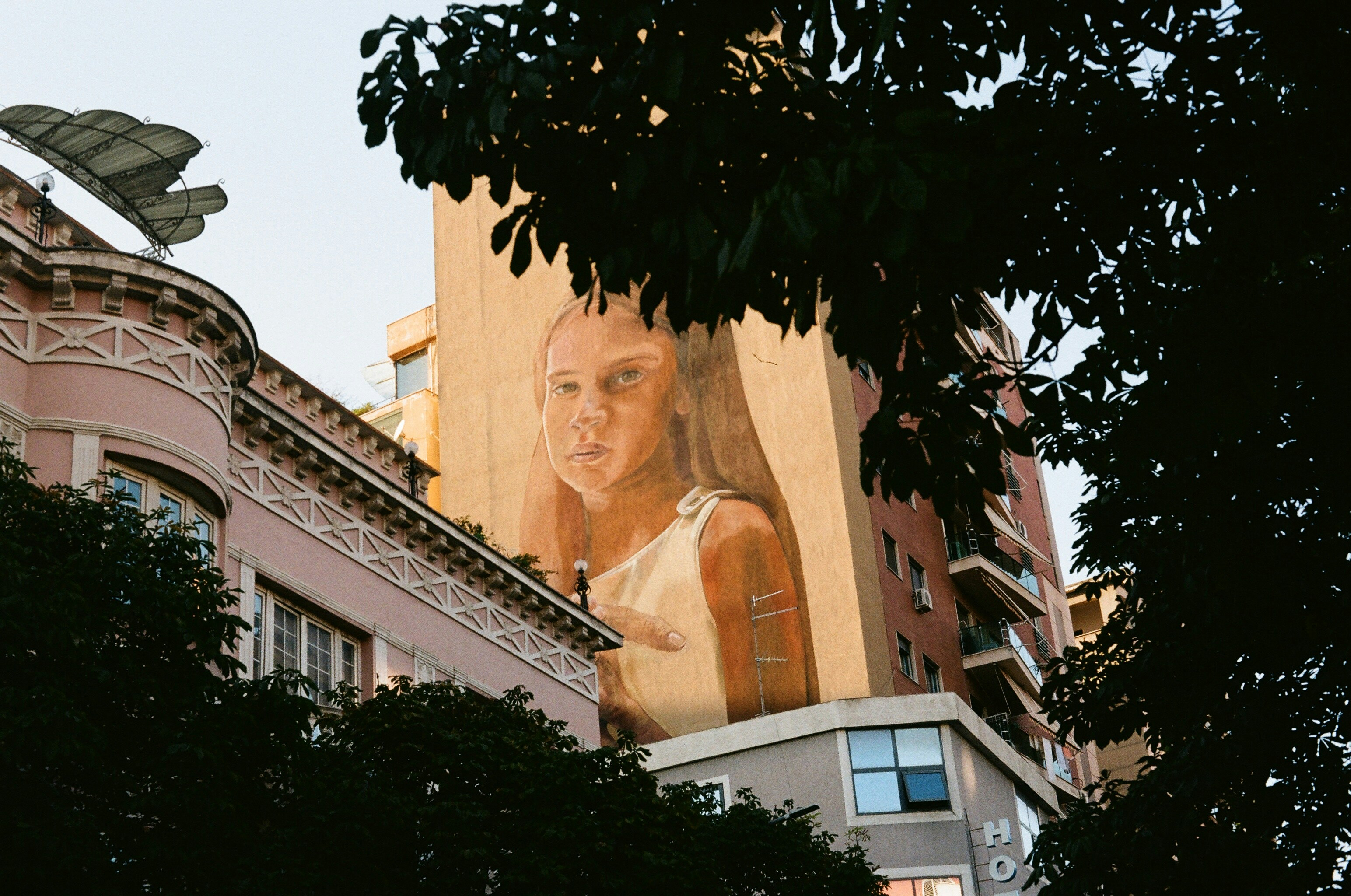 Large mural of a girl painting on an apartment building, framed by surrounding architecture and trees.