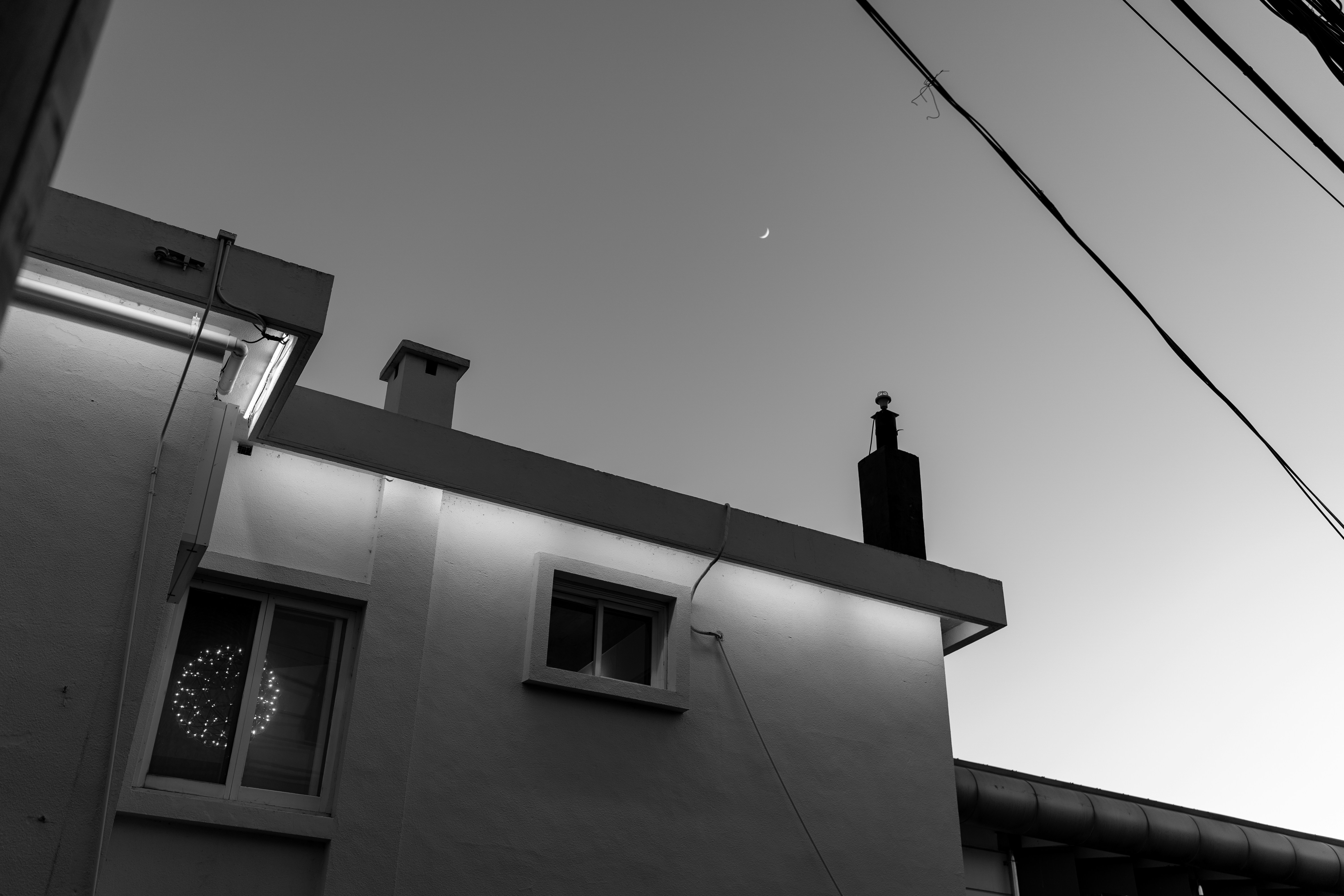 Monochrome rooftop silhouette beneath a slim crescent moon in a clear sky. Power lines cross the frame, adding geometric tension.