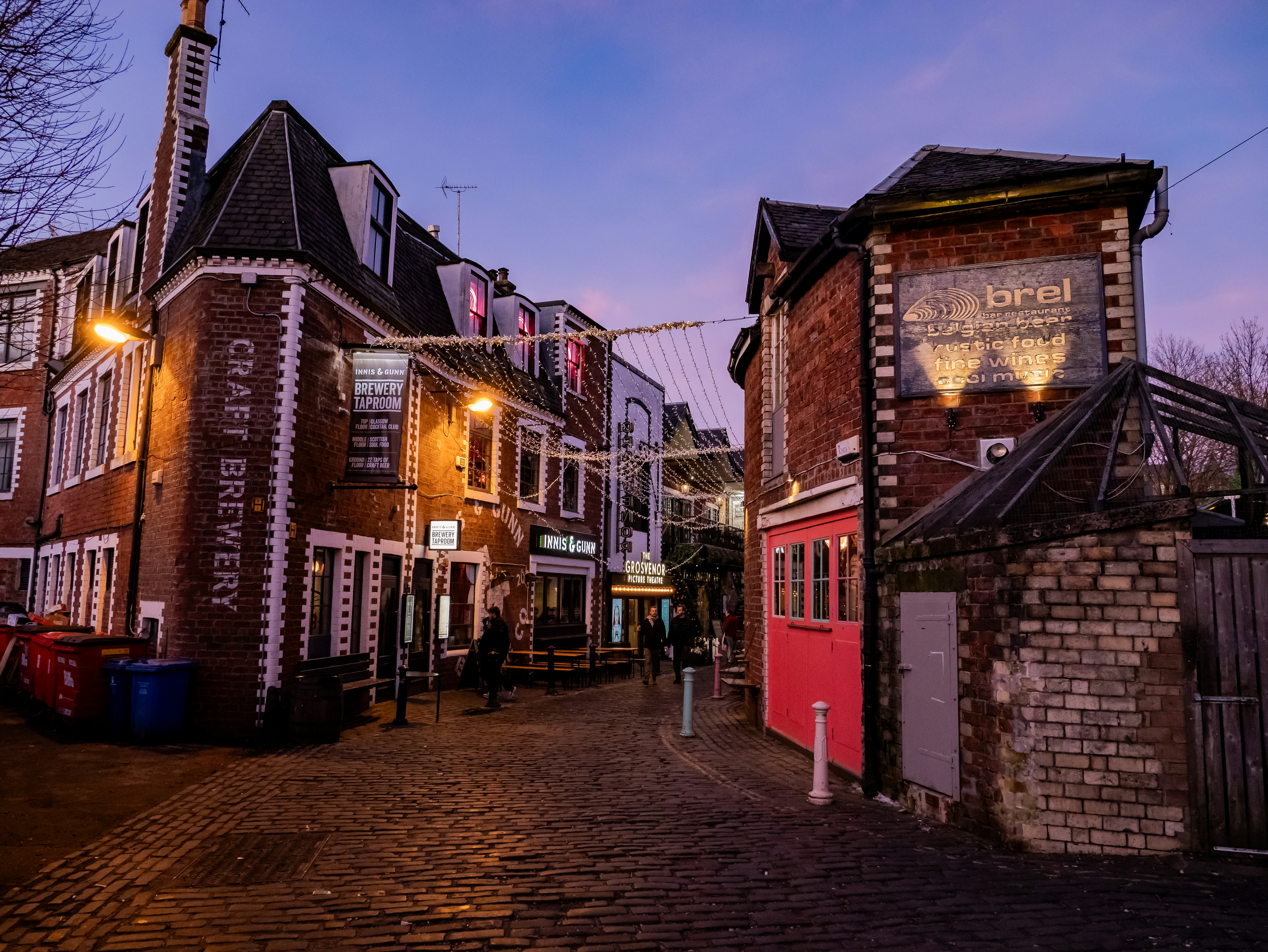 Brick-pub alley bathed in warm streetlight with string lights overhead and a cobblestone street. A cozy, historic lanescape lined with pubs and signs.